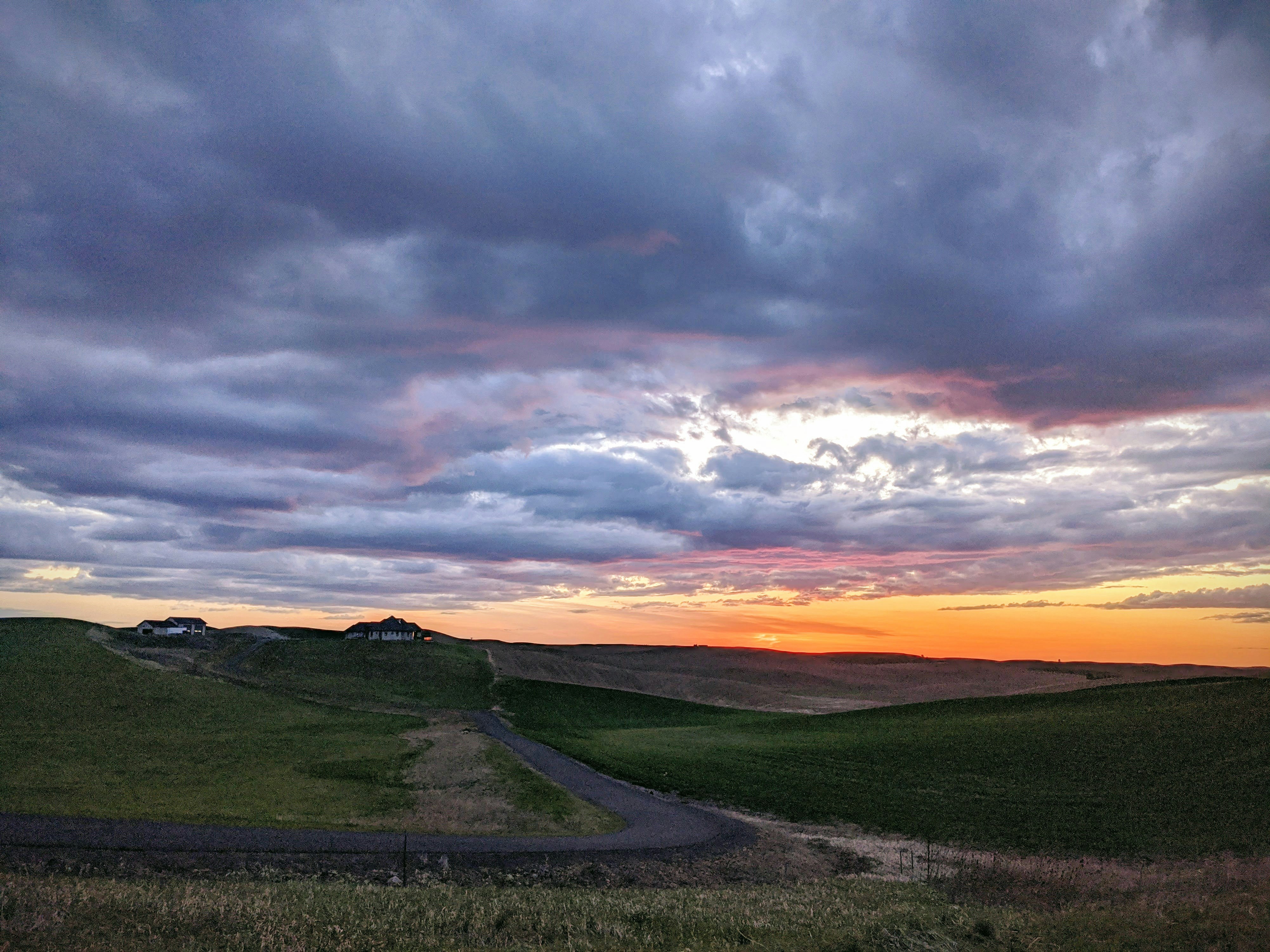 Vibrant sunset illuminating rolling hills under a dramatic sky filled with clouds. A winding road leads through the lush landscape.