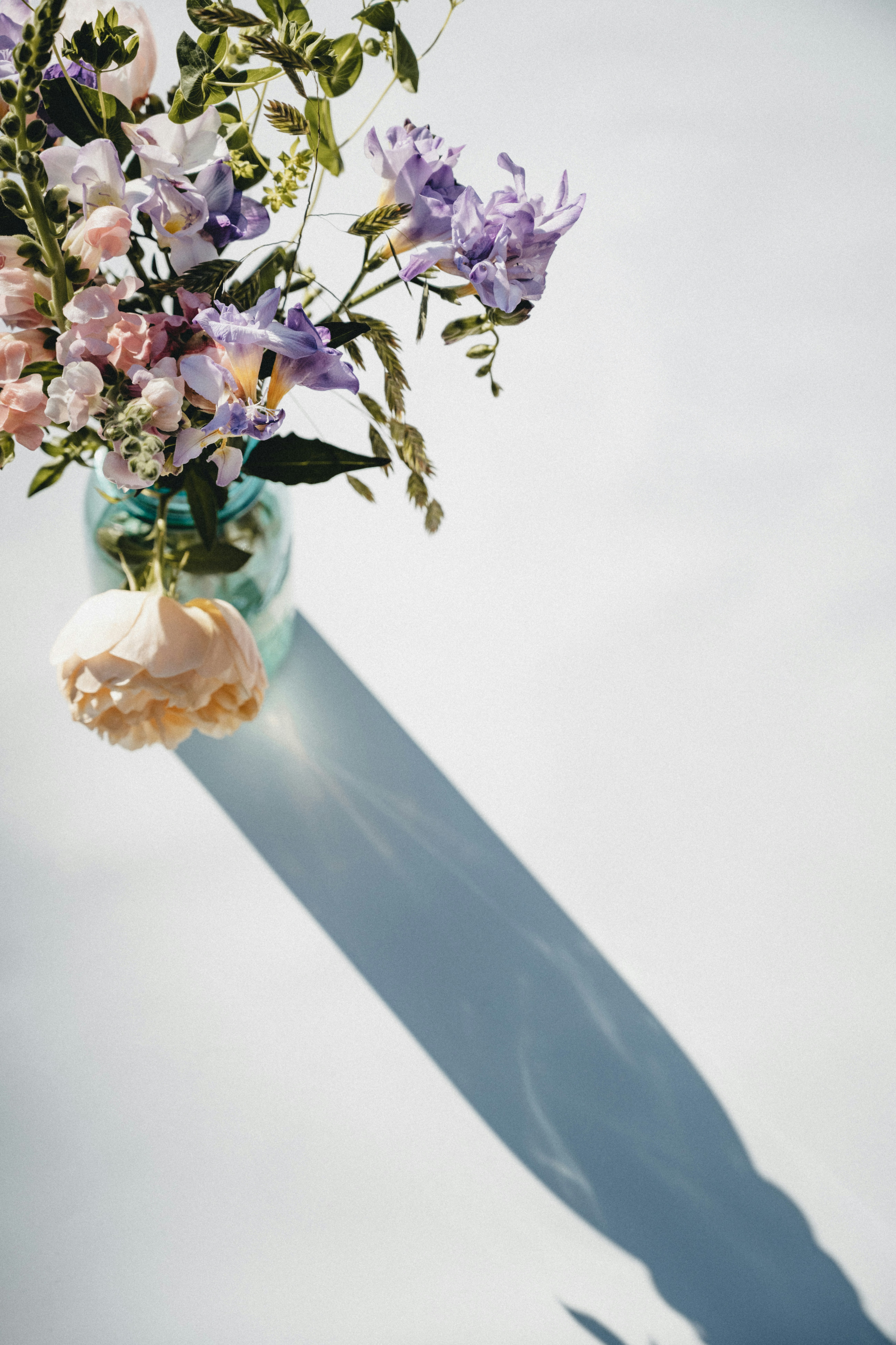 Pastel bouquet in a glass vase photographed on a bright surface, with a long diagonal shadow stretching across the scene.