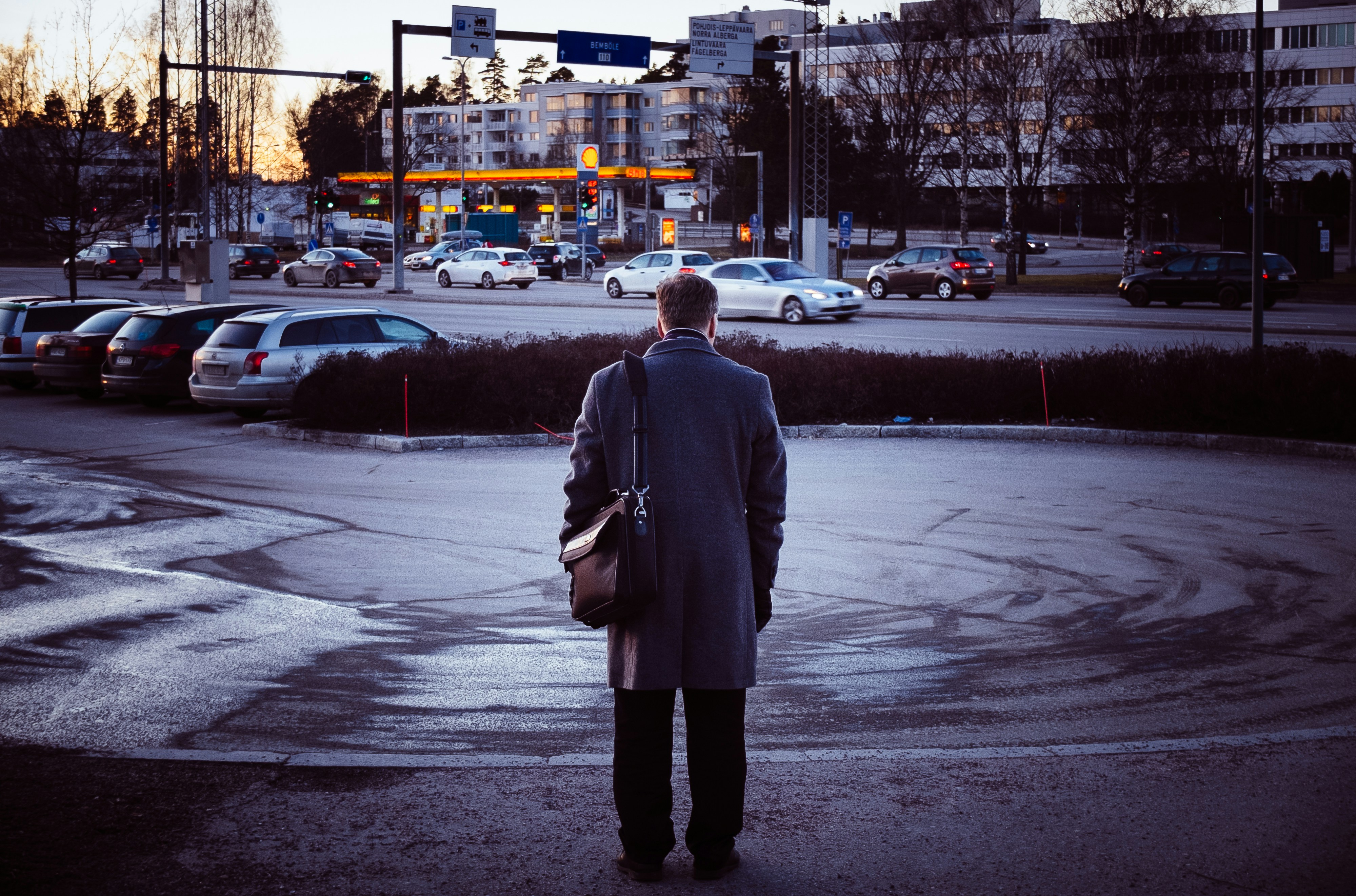 Solitary figure with a shoulder bag stands in a dim parking lot at dusk, facing a busy street where city lights are just turning on. The scene emphasizes solitude amid urban life.