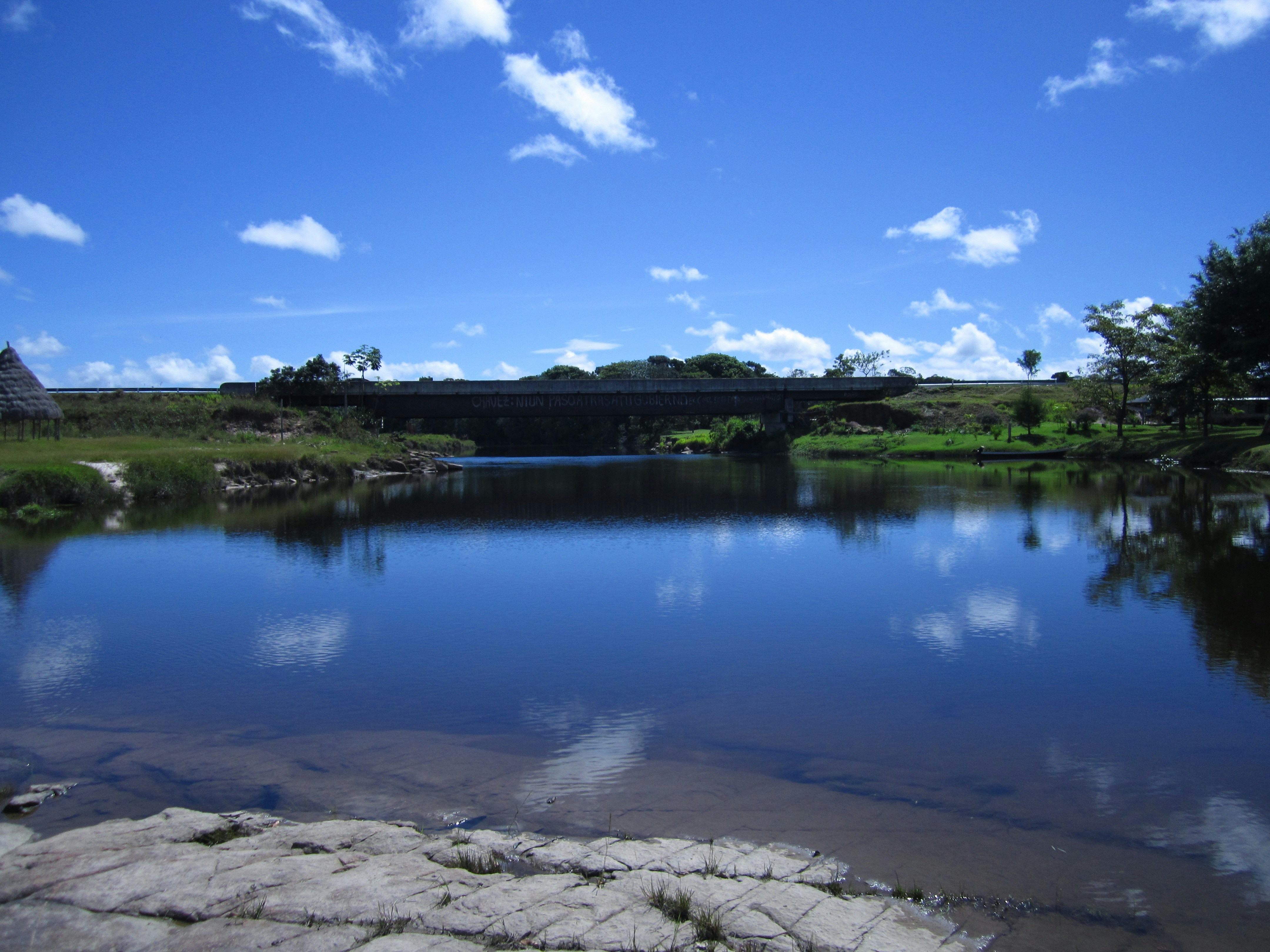 Calm river reflecting blue sky and scattered clouds, bordered by lush greenery and rocky foreground.