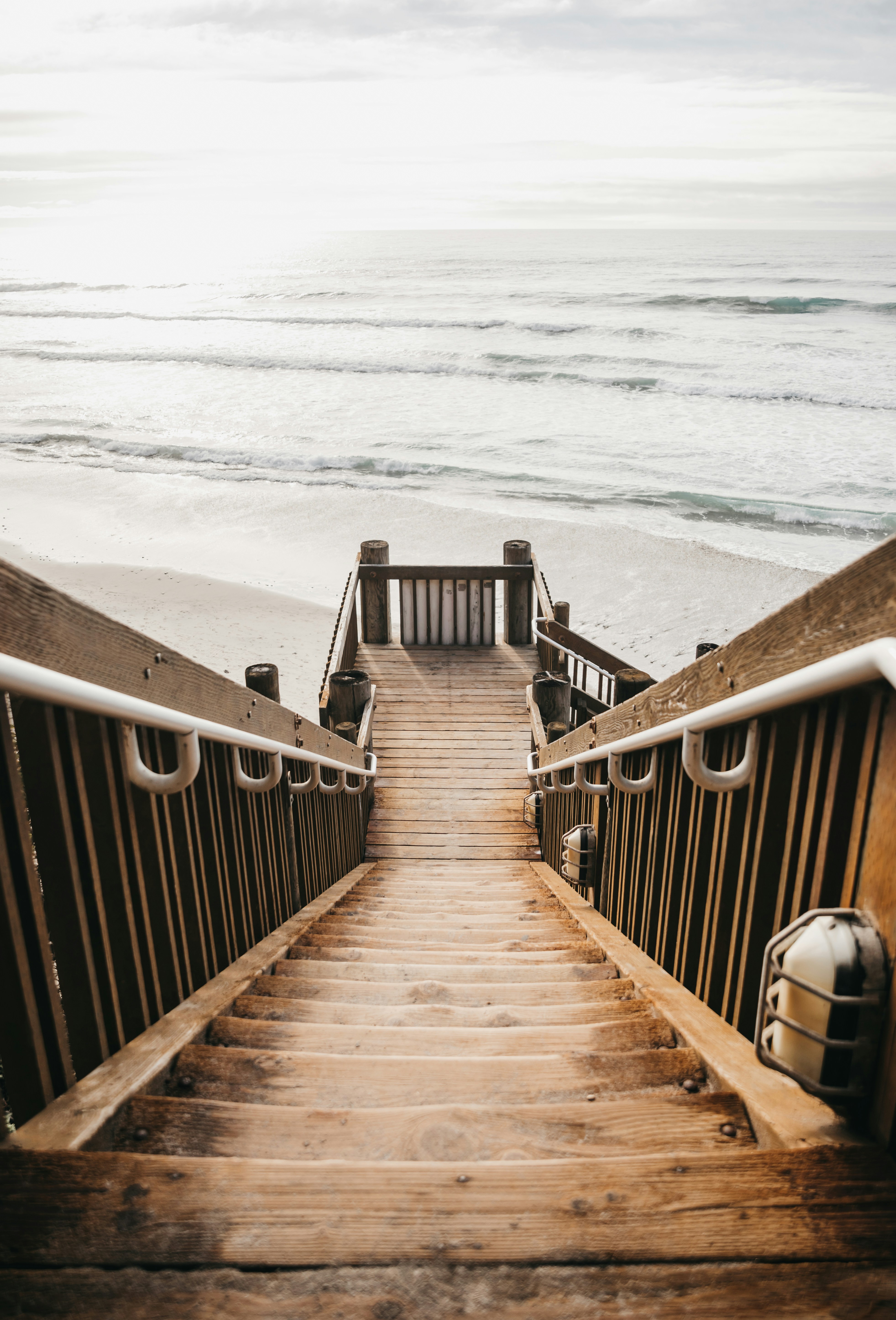 Wooden staircase leading down to a sandy beach, framed by gentle ocean waves and a soft sky. The scene evokes a sense of tranquility.