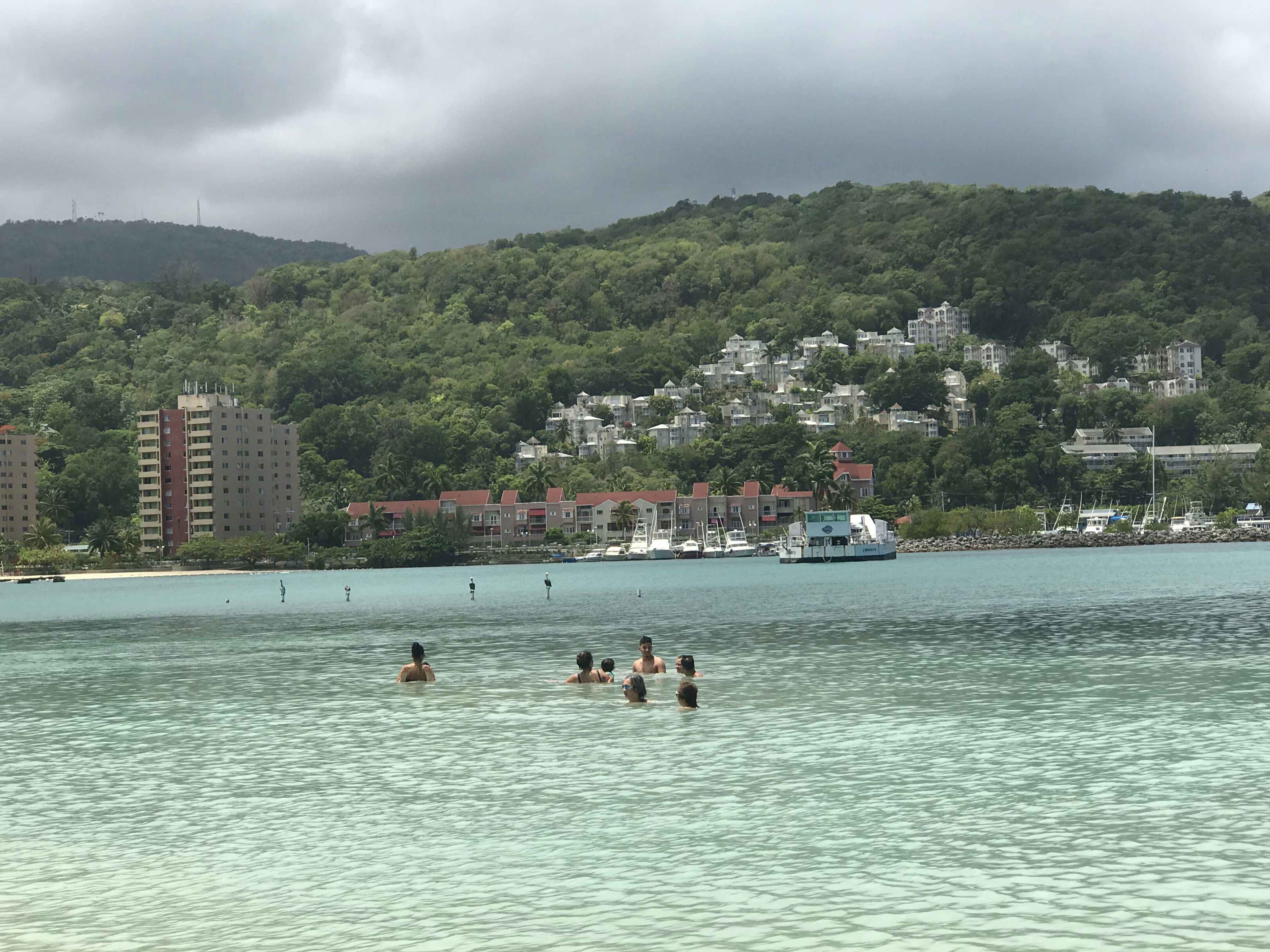 Group of swimmers enjoying the calm waters near a coastal resort, with green hills and buildings in the background.
