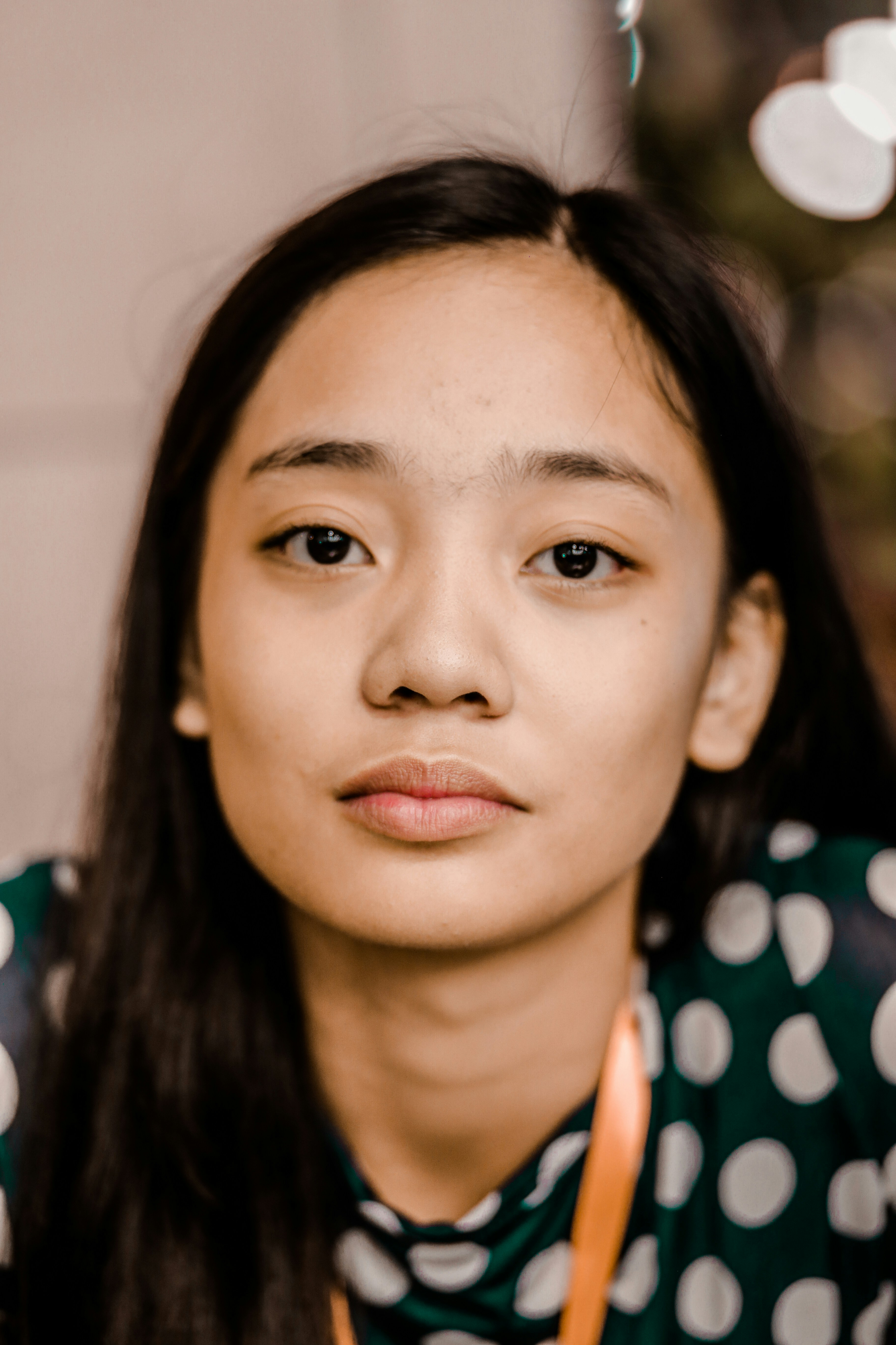A young woman with a calm expression gazes directly at the viewer, adorned in a polka-dotted outfit. Soft bokeh lights create a warm atmosphere.