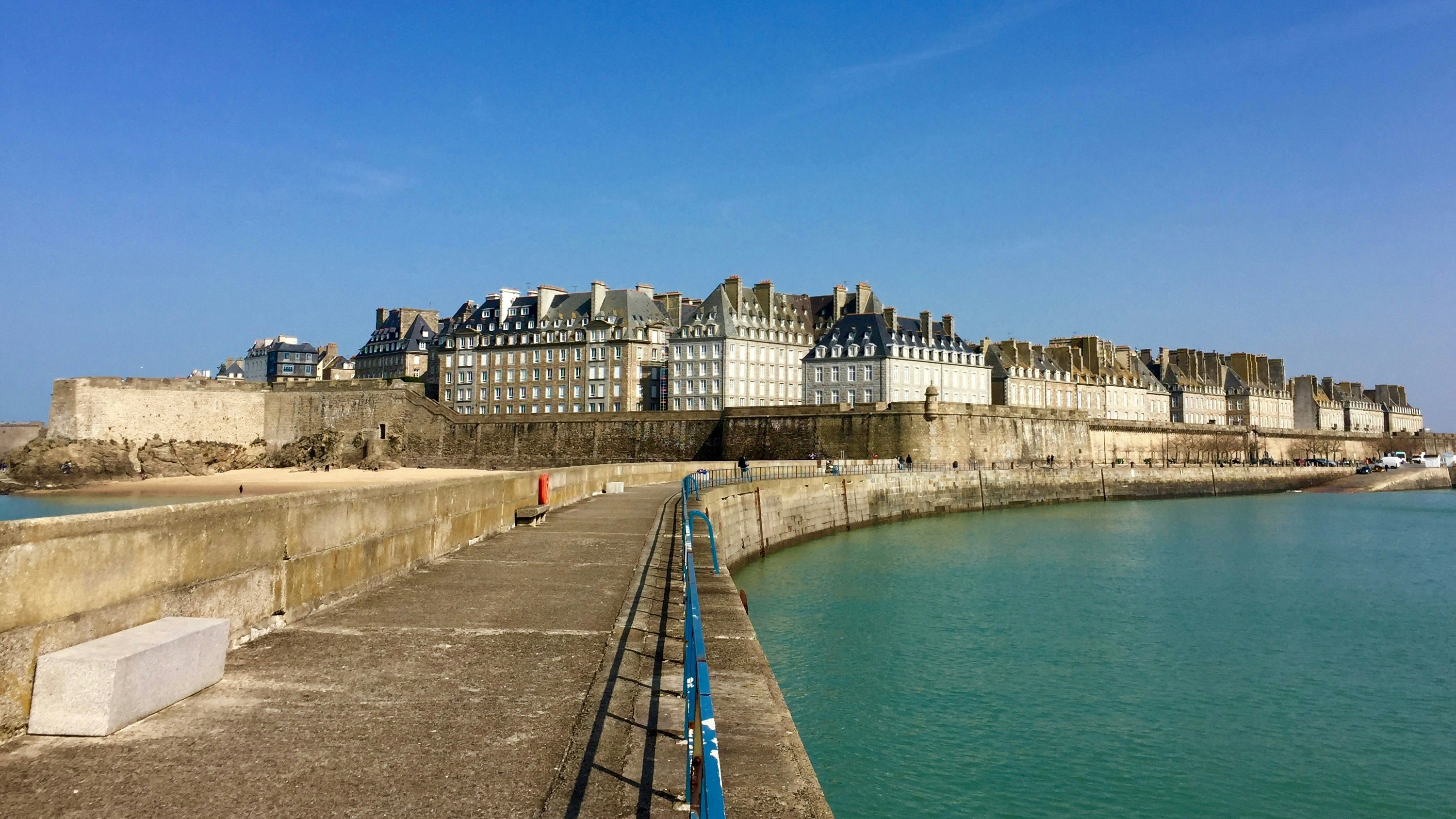 Historic buildings line the fortified walls of Saint-Malo, overlooking the calm turquoise waters. A serene promenade leads towards the ancient structures.