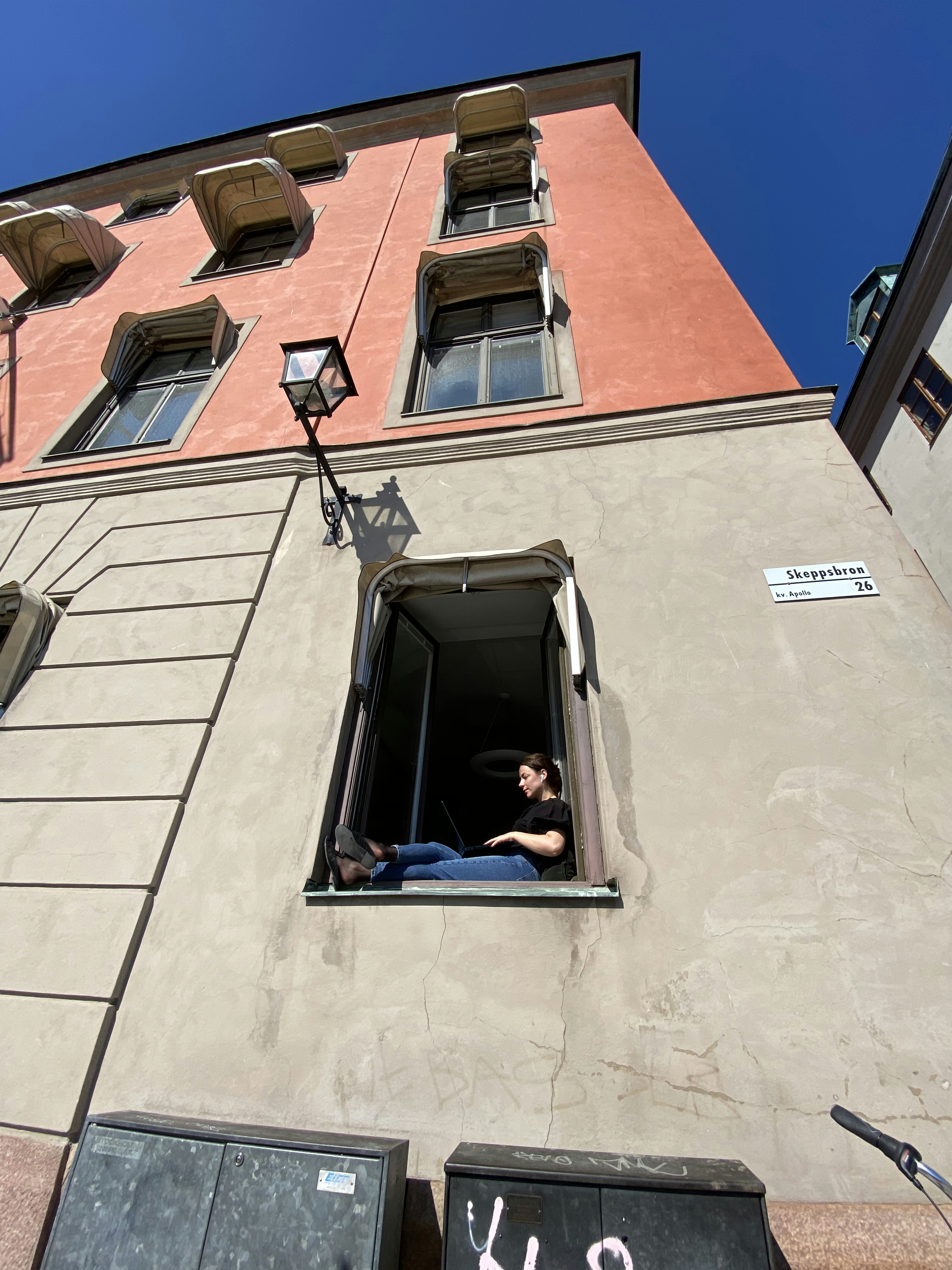 A person sits comfortably in a window, absorbed in their laptop, framed by a colorful building facade. The scene captures a blend of urban life and personal tranquility.