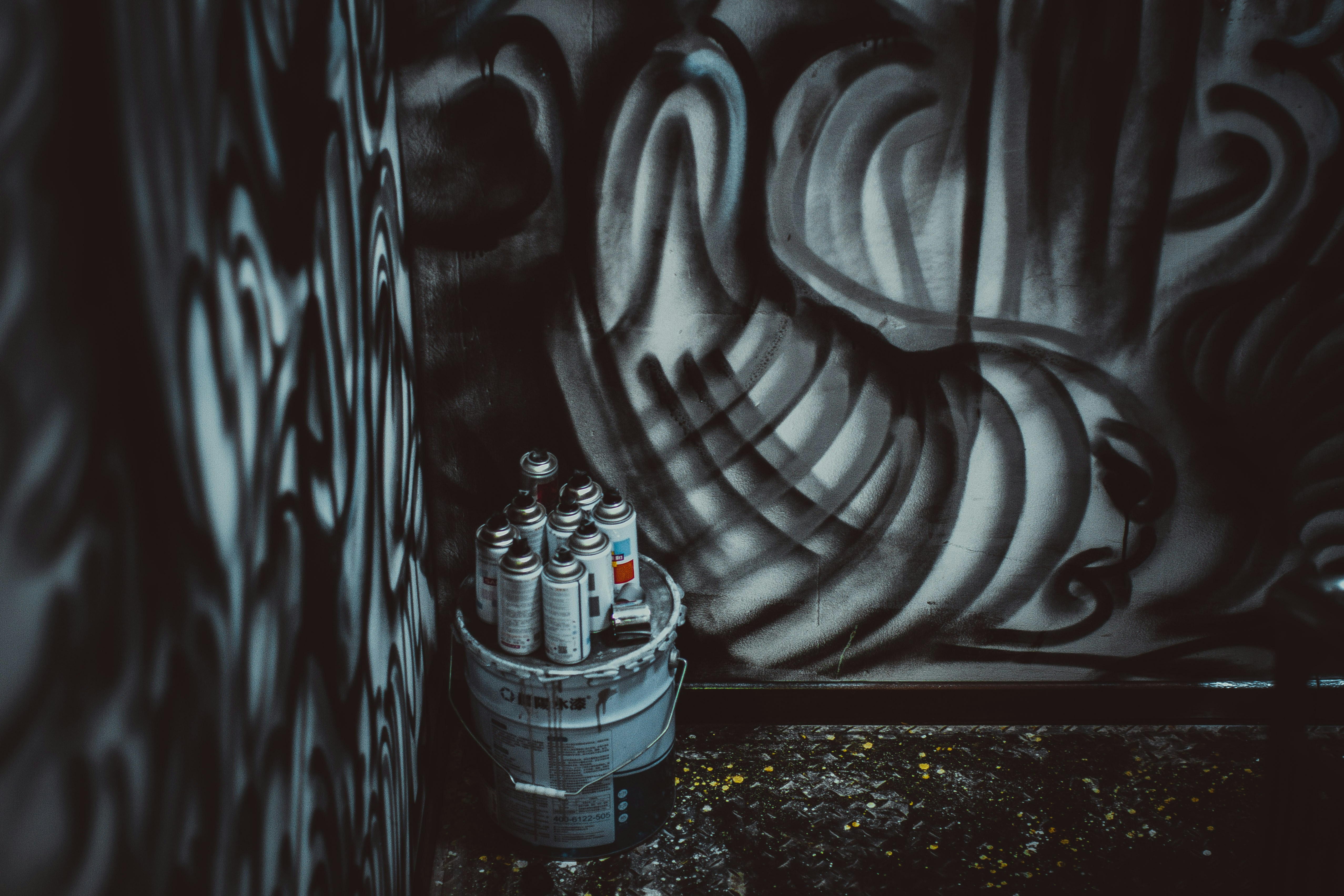 A collection of spray paint cans rests in a bucket against a backdrop of intricate graffiti patterns. The scene captures the vibrant spirit of urban creativity.