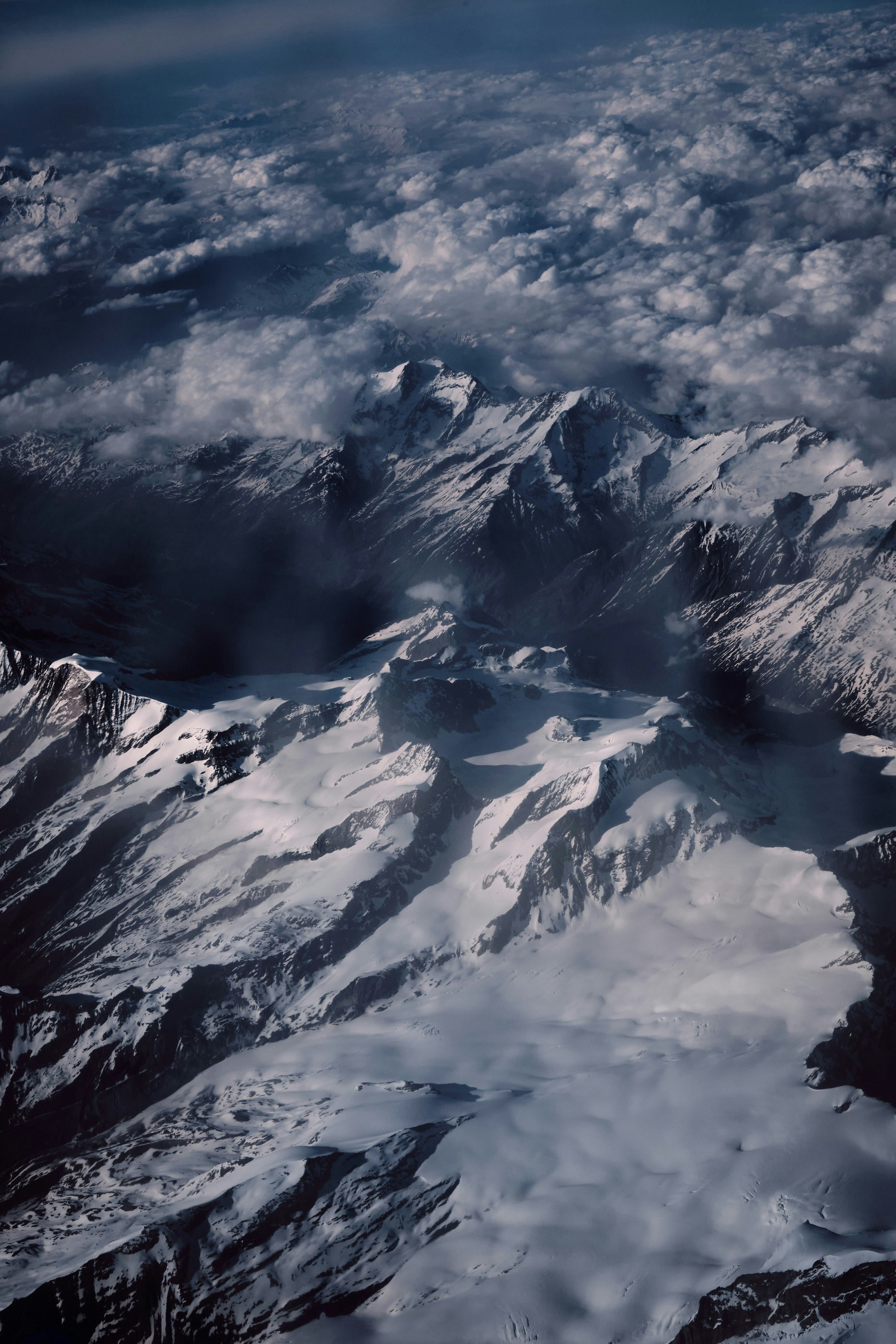 Aerial view of snow-capped mountains and glaciers beneath a blanket of clouds, showcasing the rugged beauty of nature's high altitudes.