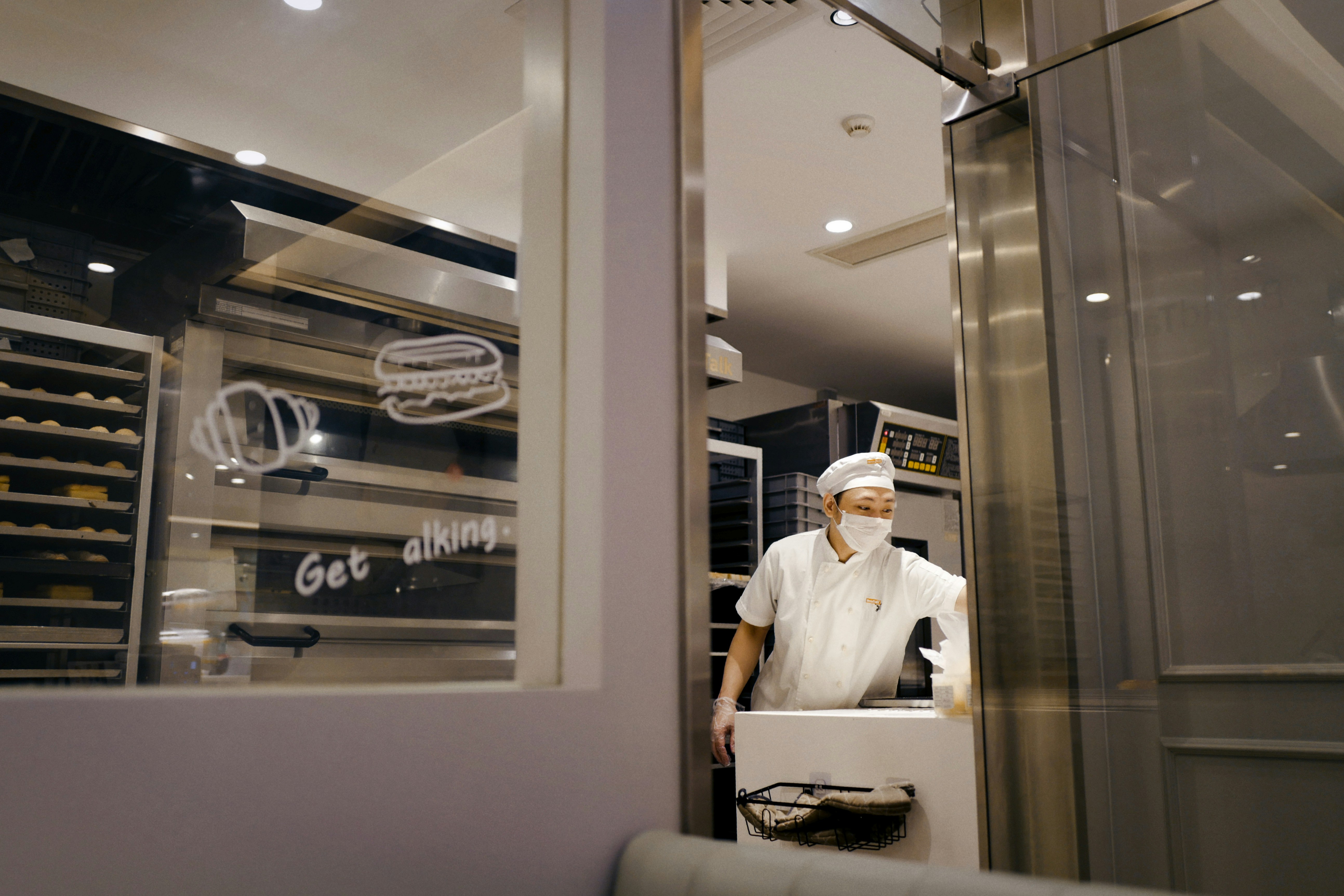 Baker in a white uniform working behind a glass partition, surrounded by baking trays and equipment. The scene captures the essence of culinary artistry.