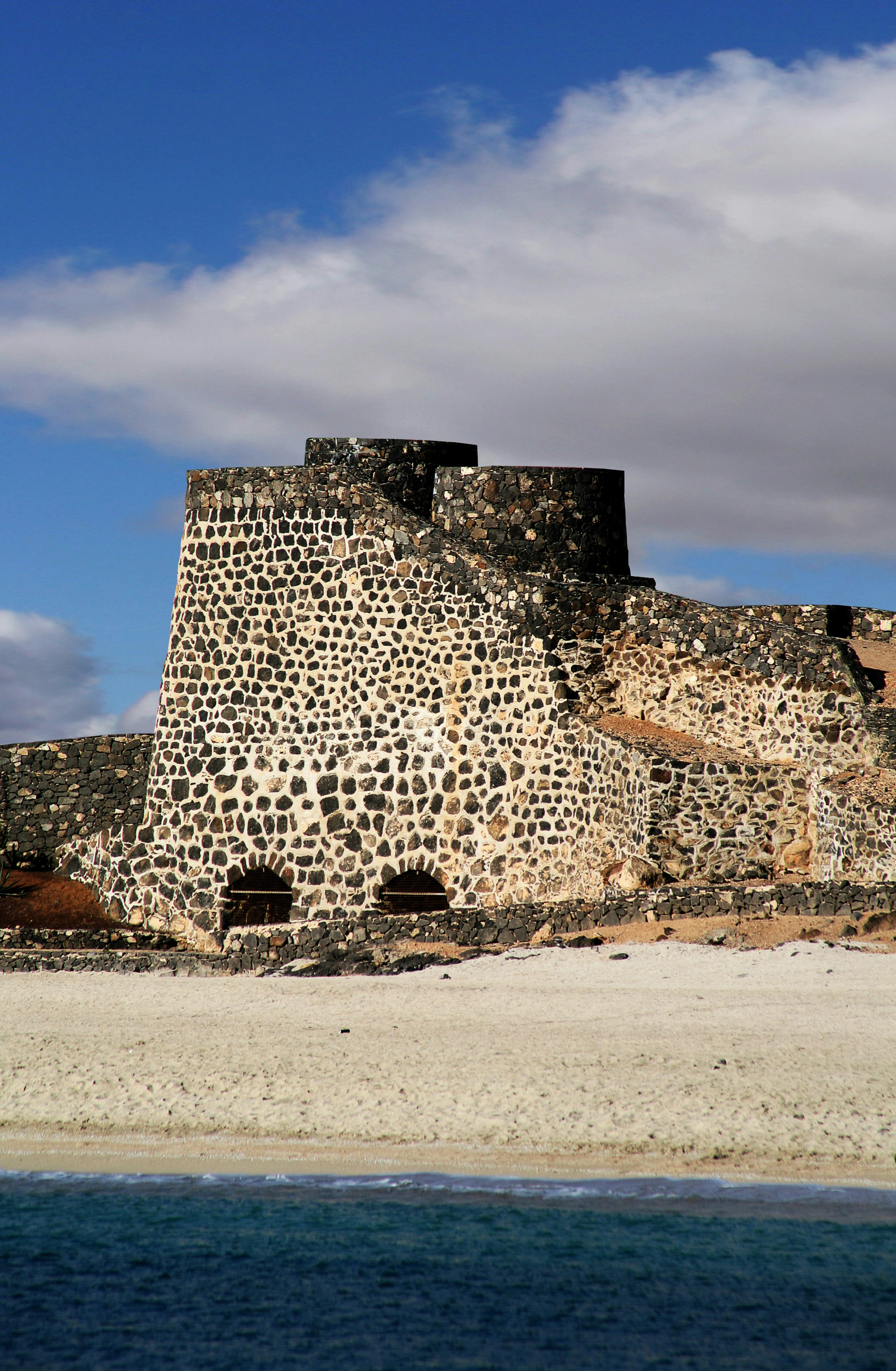Weathered stone tower remnants stand against a backdrop of sandy beach and azure waters, illustrating a historical coastal defense structure.