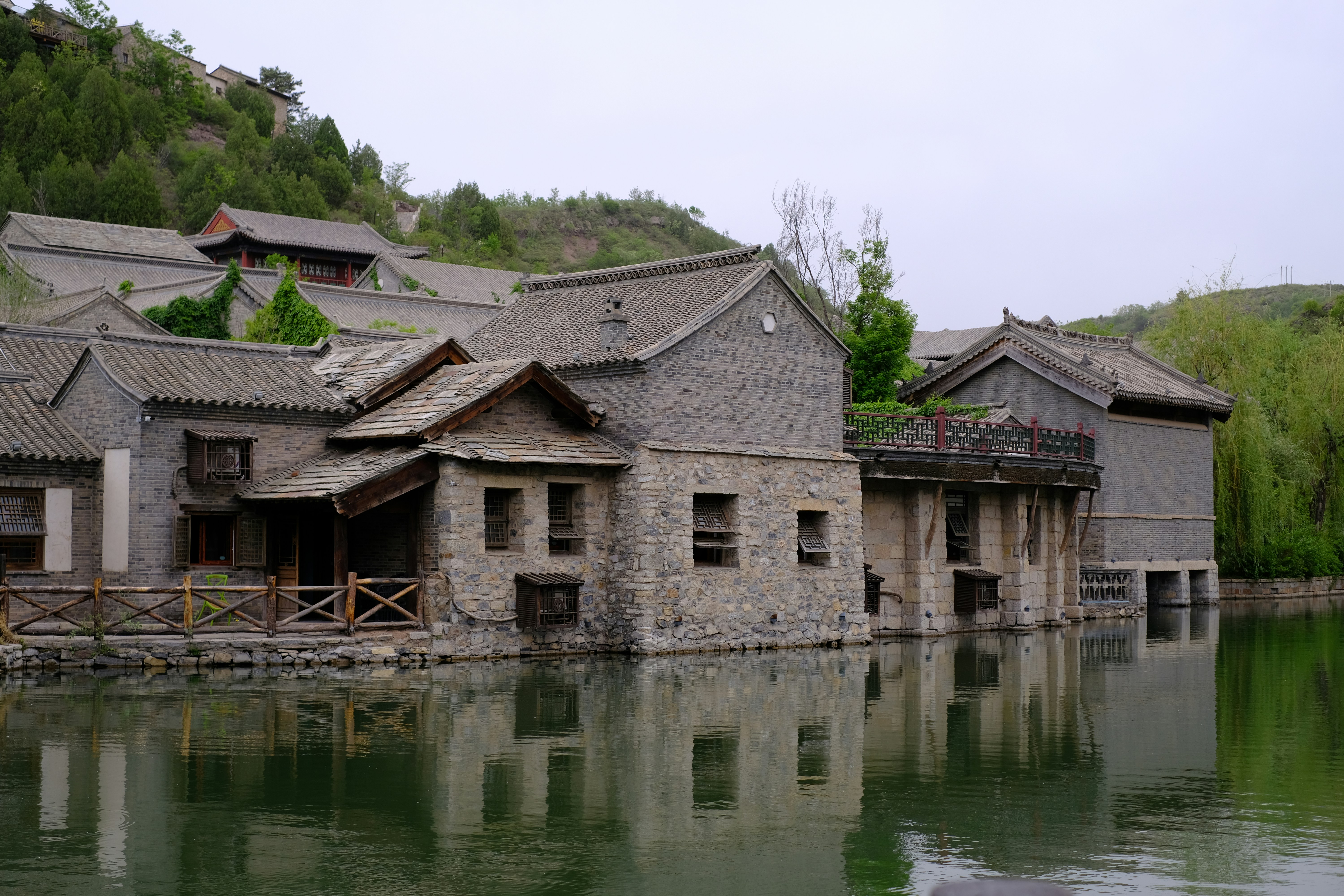 Historic stone houses line a serene lake, reflecting traditional architecture in calm waters. Lush greenery frames the scene.