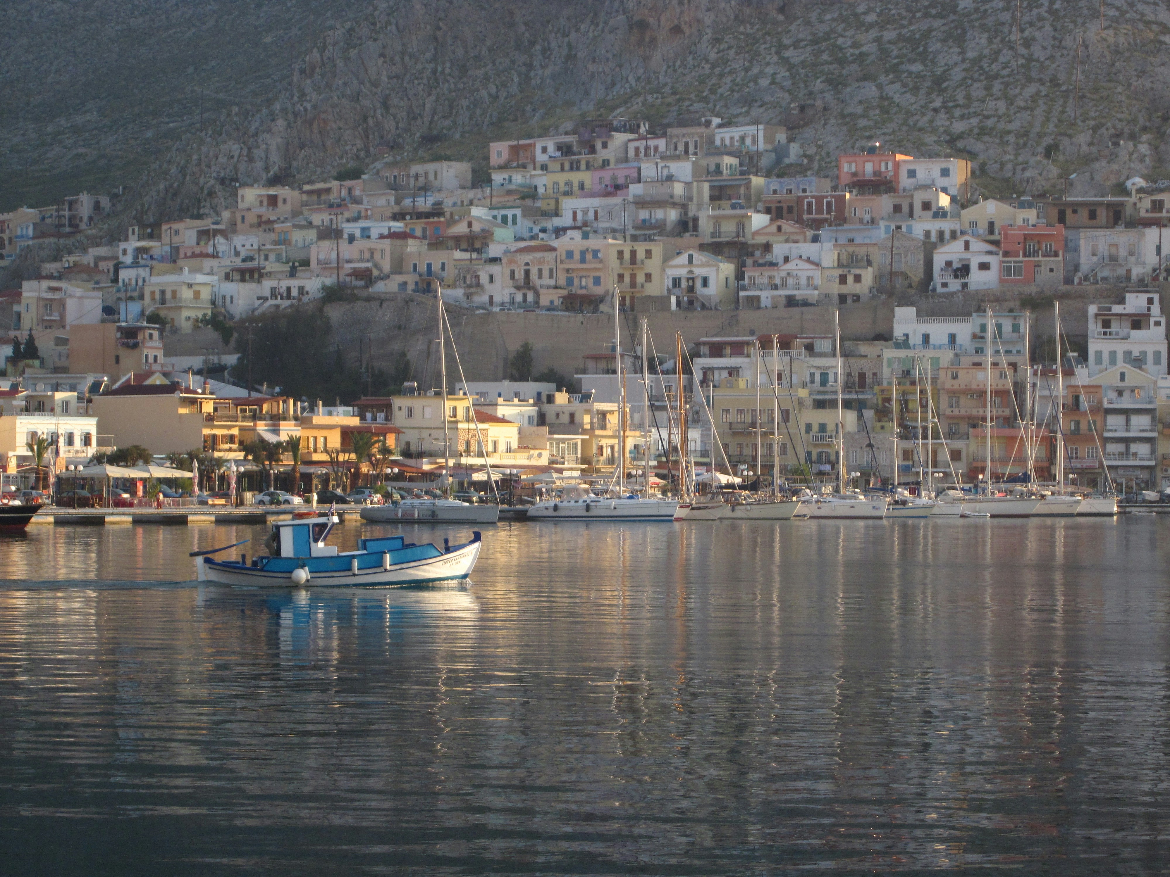 Blue fishing boat anchors in a calm harbor with pastel hillside houses and sailboats lining the quay.
