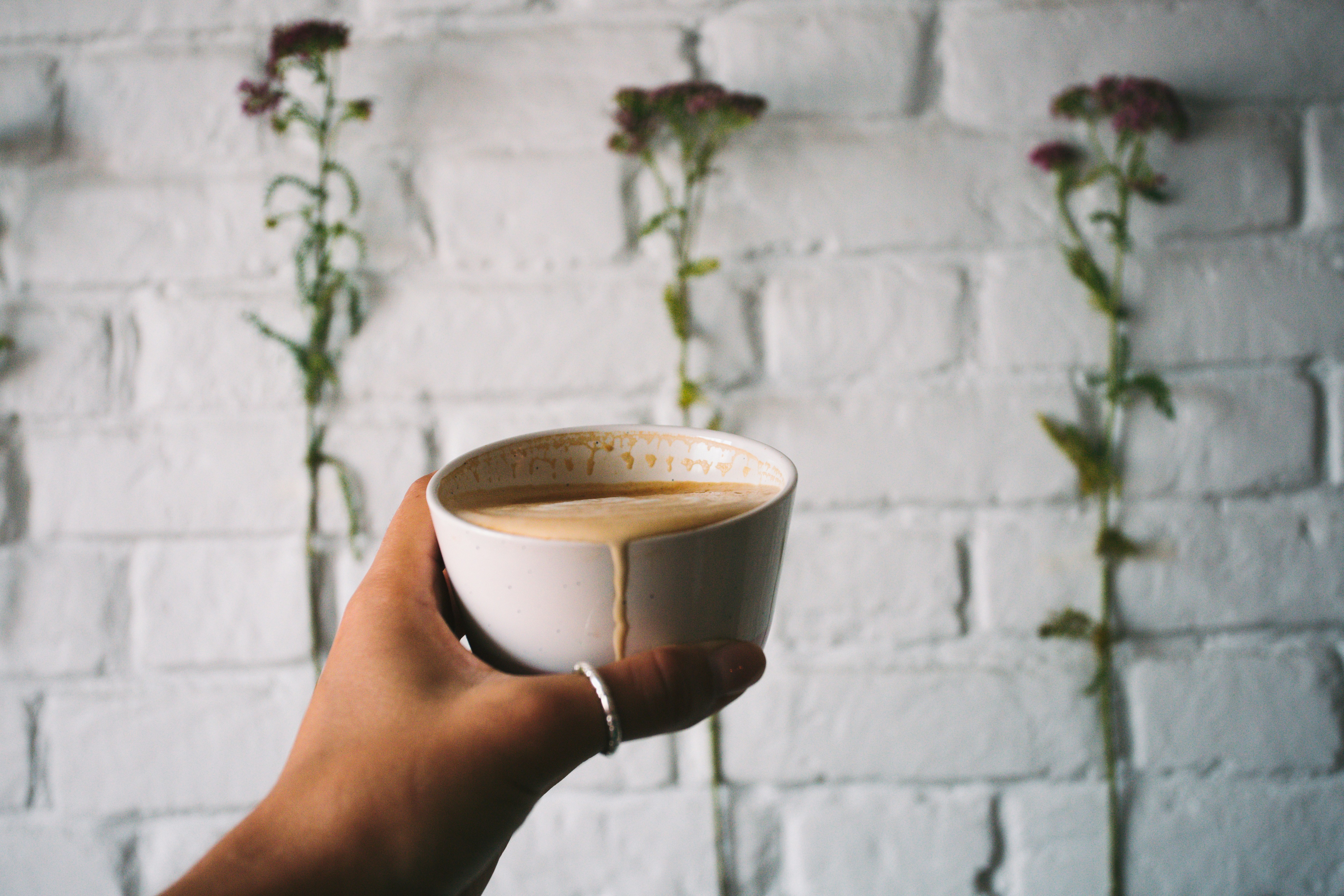 Hand holding a white cup of coffee against a textured white brick wall adorned with delicate flowers.
