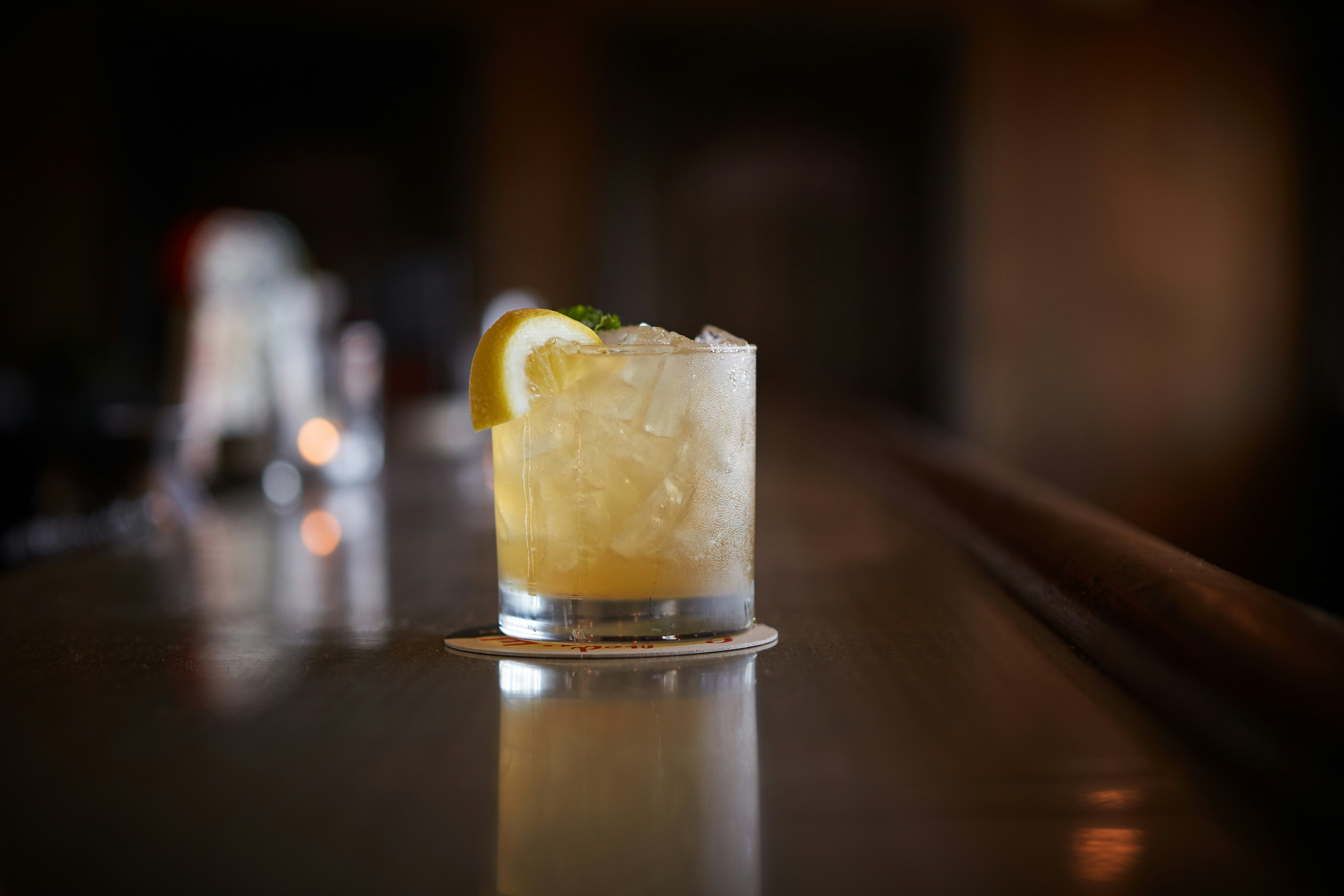 A refreshing cocktail garnished with a lemon slice, resting on a wooden bar top with blurred background elements suggesting a lively atmosphere.
