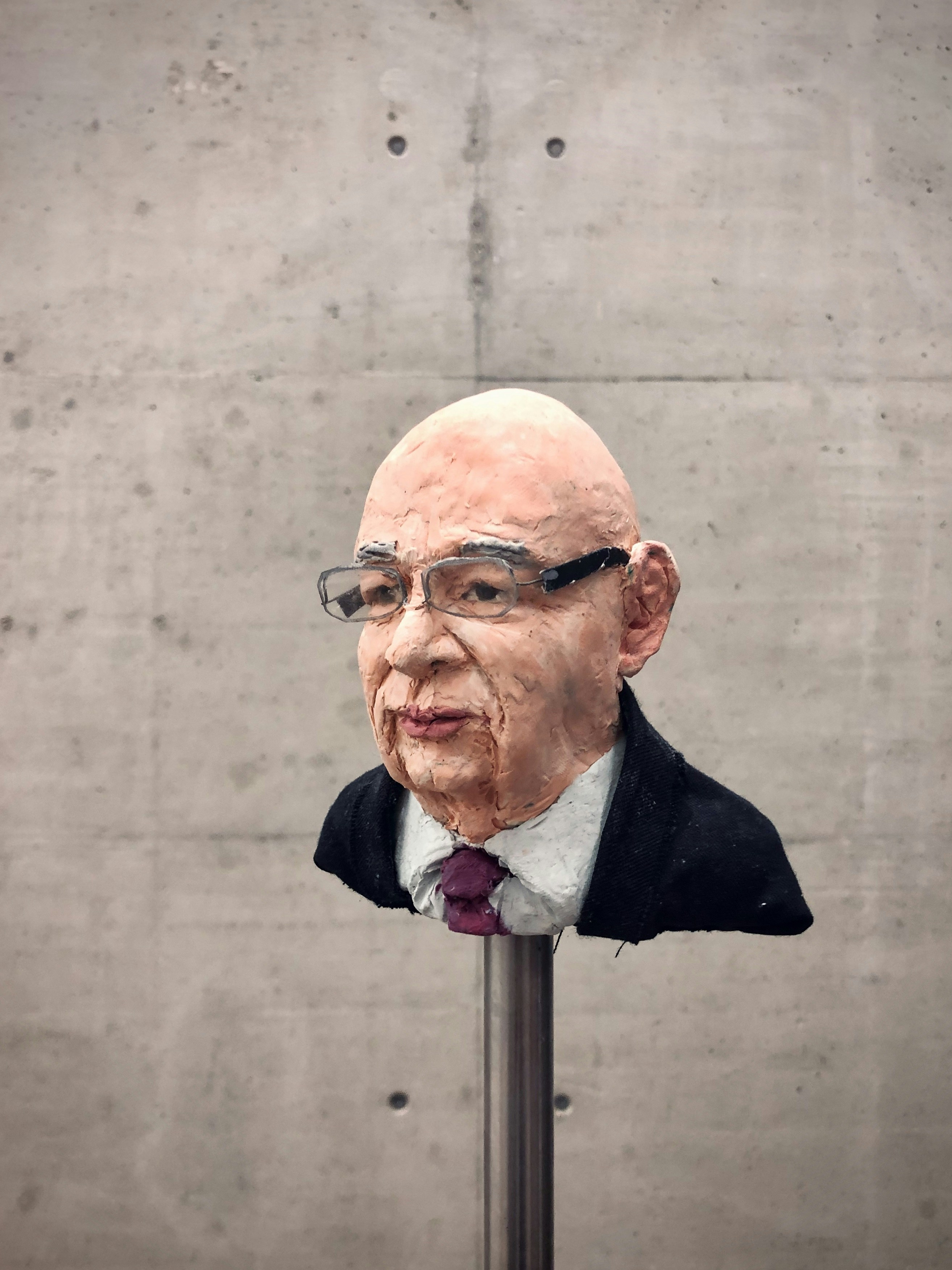 Detailed sculpture of a bespectacled man in a suit, showcasing intricate facial features and expressions against a minimalist concrete backdrop.