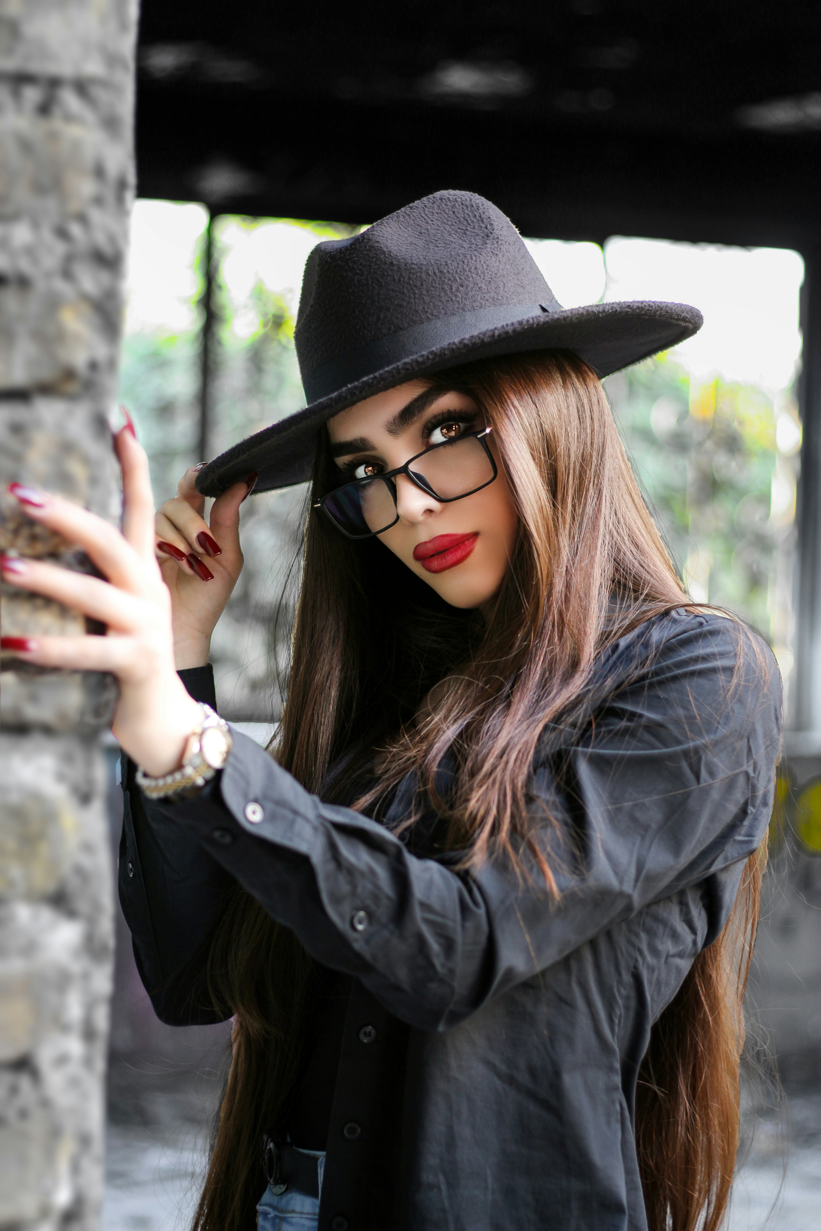 A stylish woman in a black shirt and wide-brimmed hat poses against a textured stone wall, exuding confidence and allure.