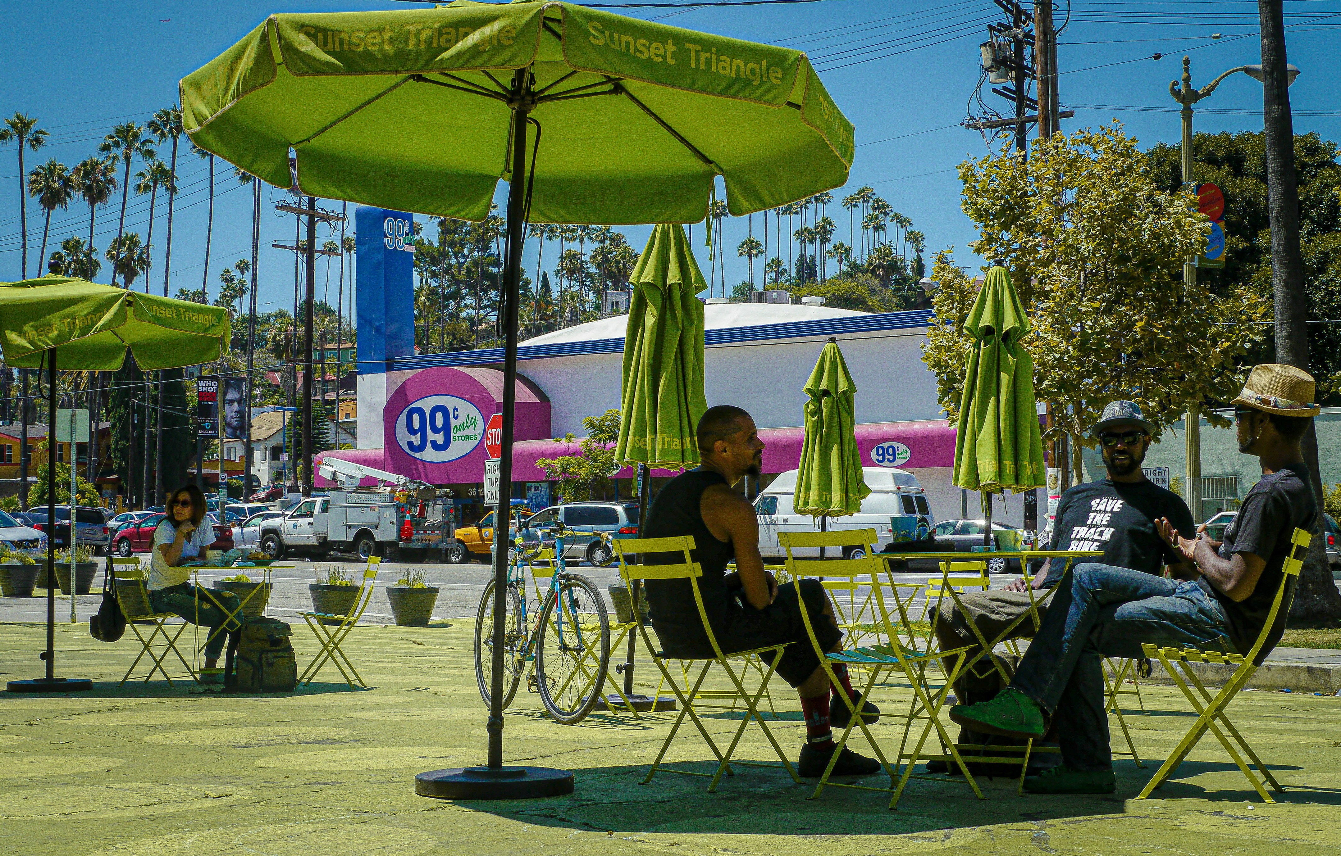 Three individuals engage in conversation at a vibrant outdoor café, surrounded by bright green umbrellas and a lively urban backdrop.