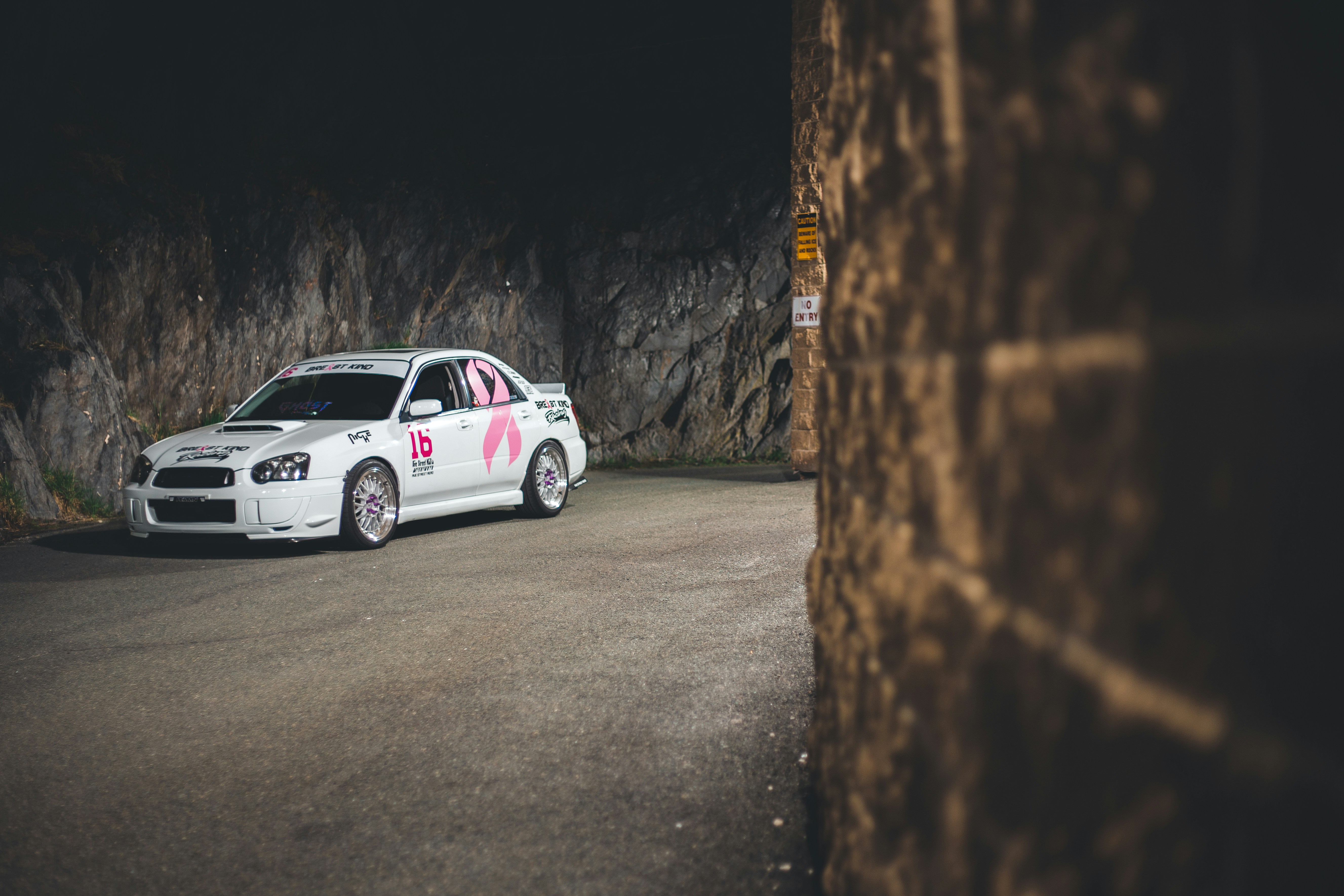 Modified white car with pink accents parked in a dimly lit urban setting, surrounded by rugged stone walls.