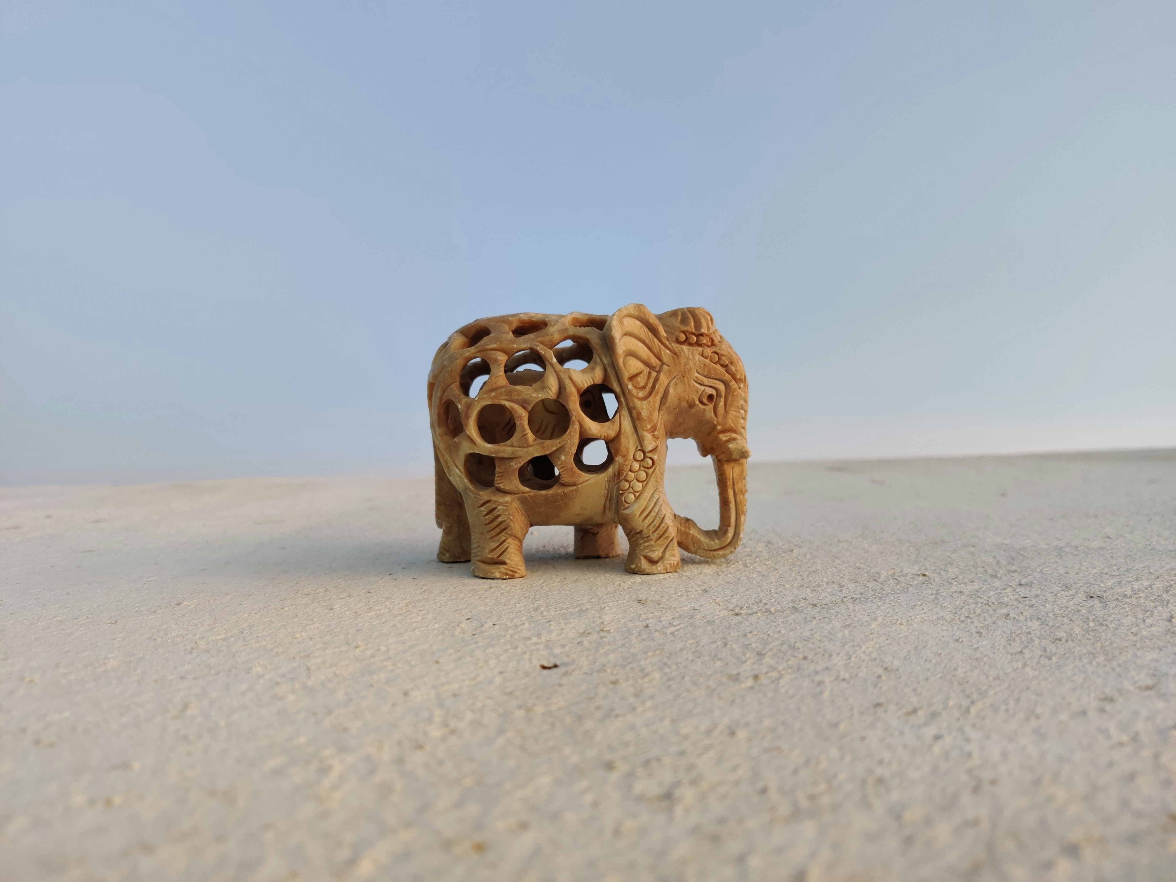 Small carved elephant sculpture with geometric holes sits on sandy beach under a clear blue sky.