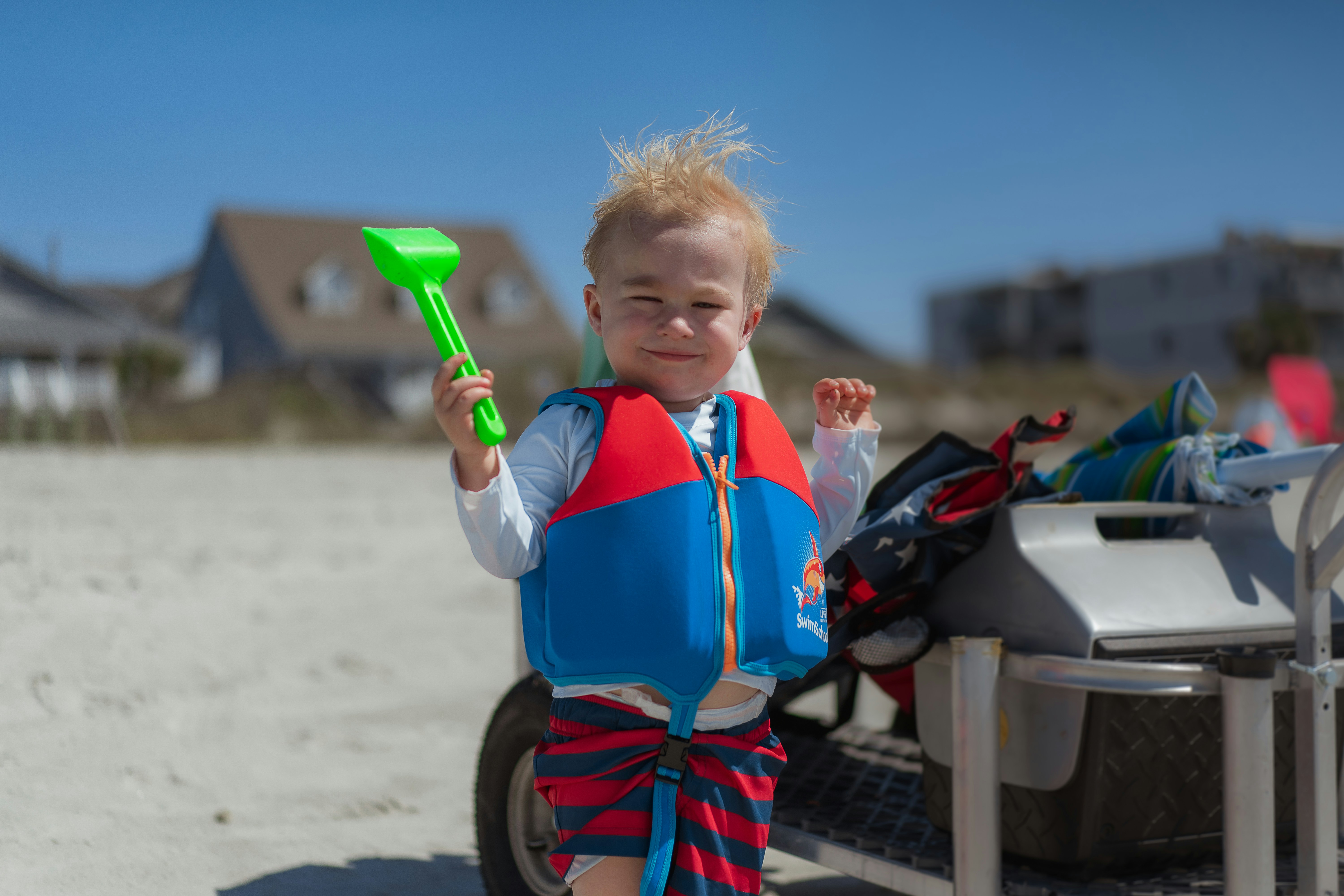 Young child in a colorful life jacket holding a green shovel, smiling on a sandy beach with beach gear in the background.