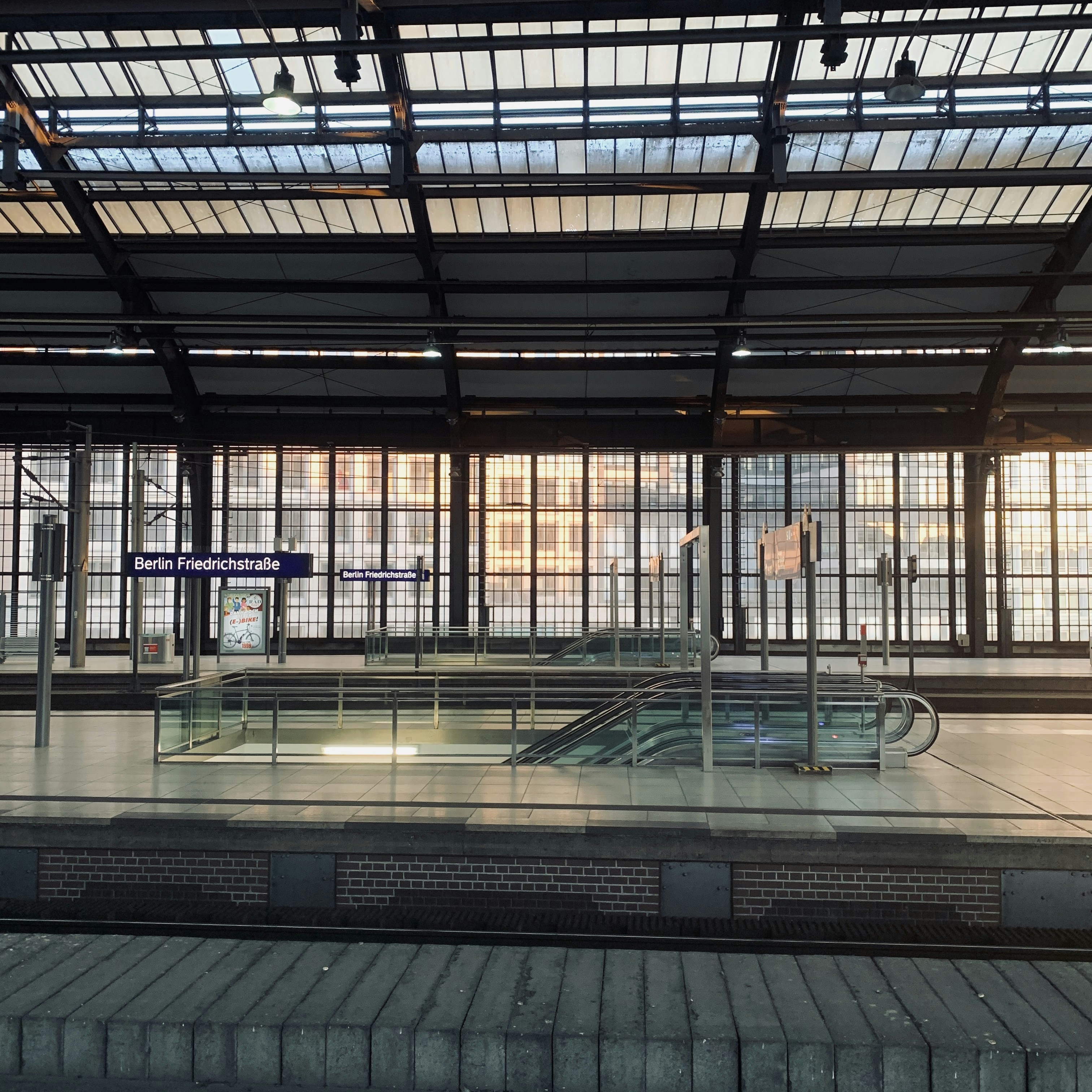 Modern train station platform featuring escalators and large glass windows, bathed in soft morning light.