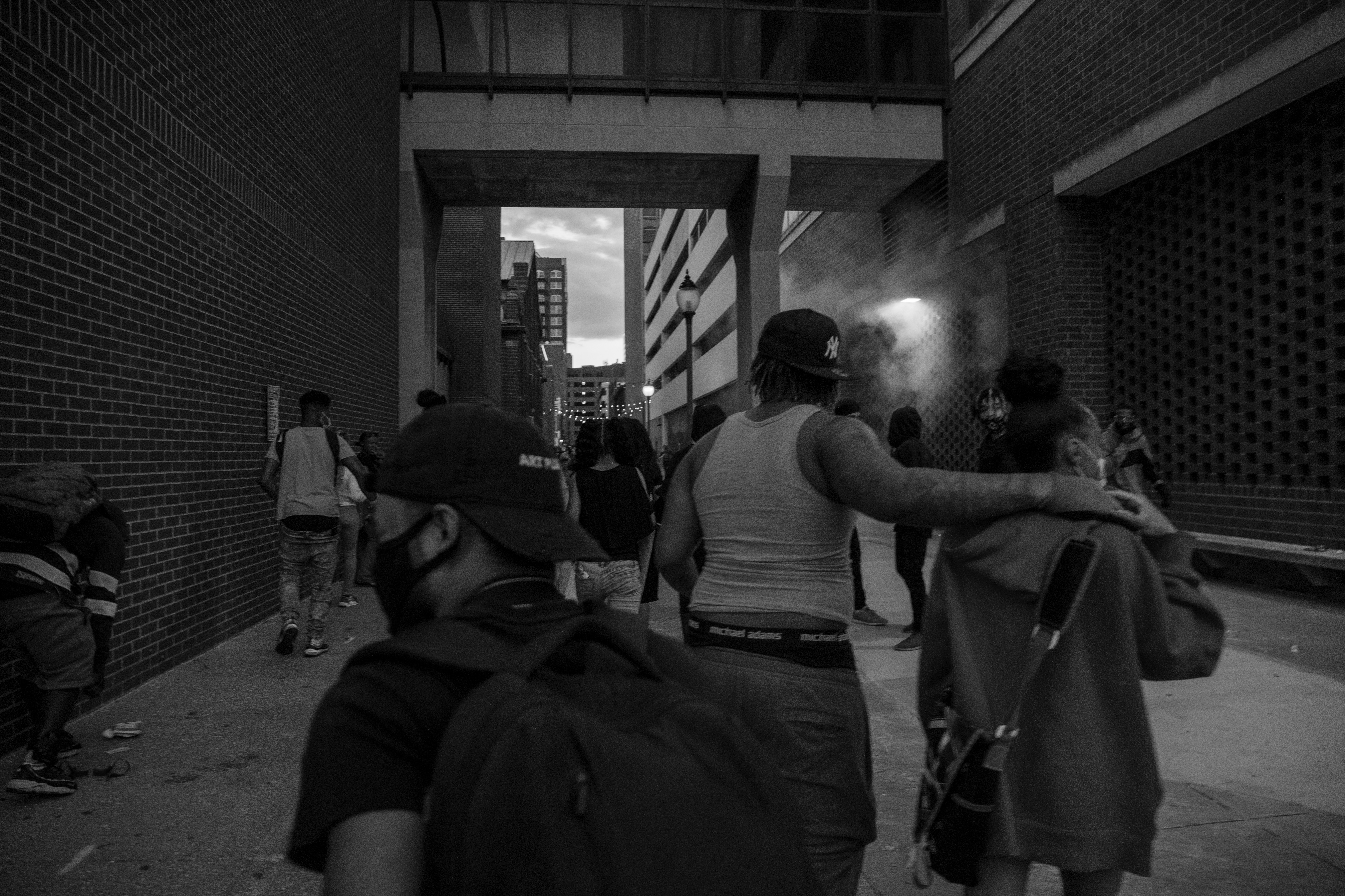 Protesters navigate a narrow alley filled with tear gas, seeking safety amidst urban buildings.