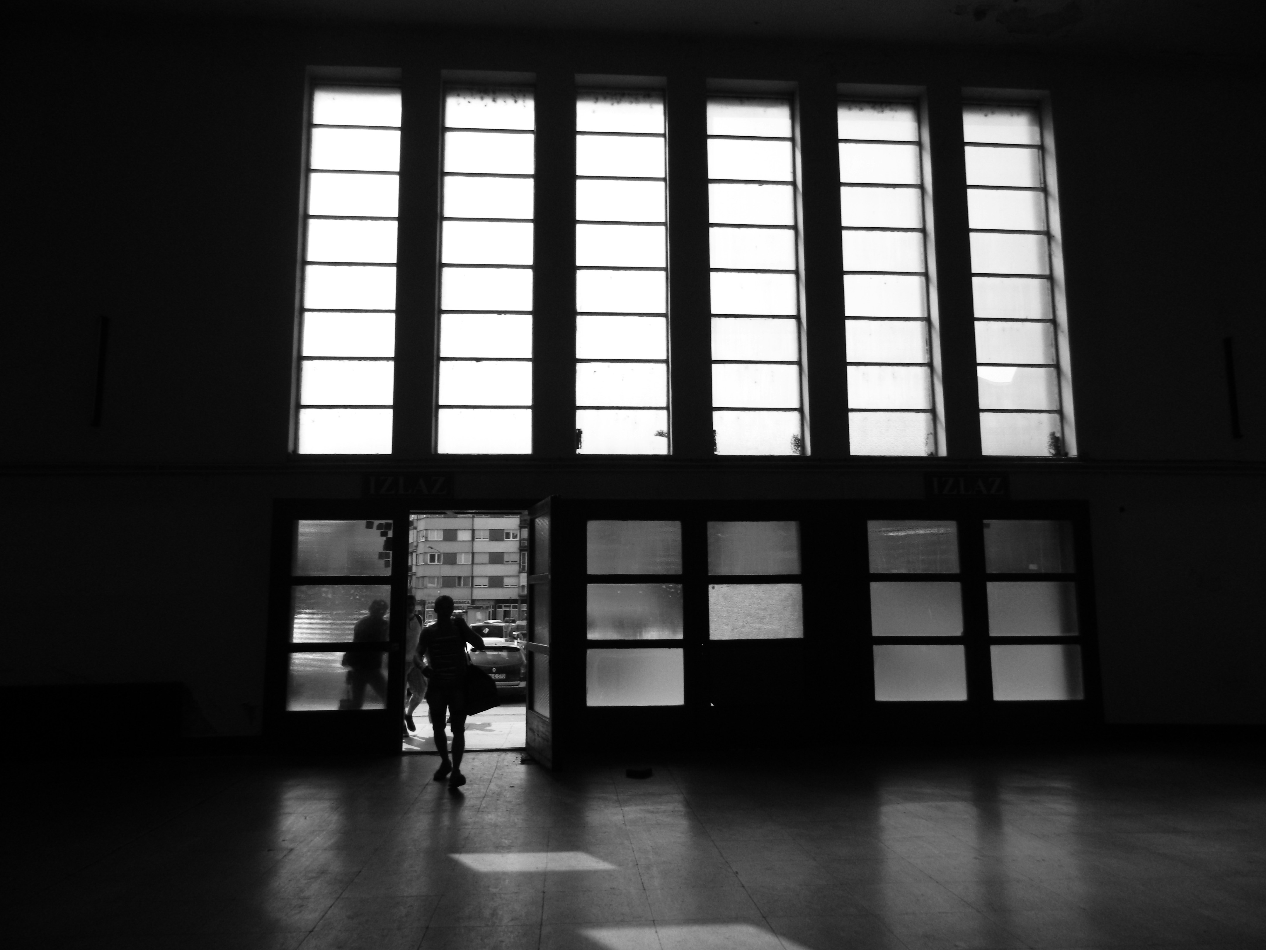 Silhouetted figures walk through a large windowed entrance in a dimly lit station.