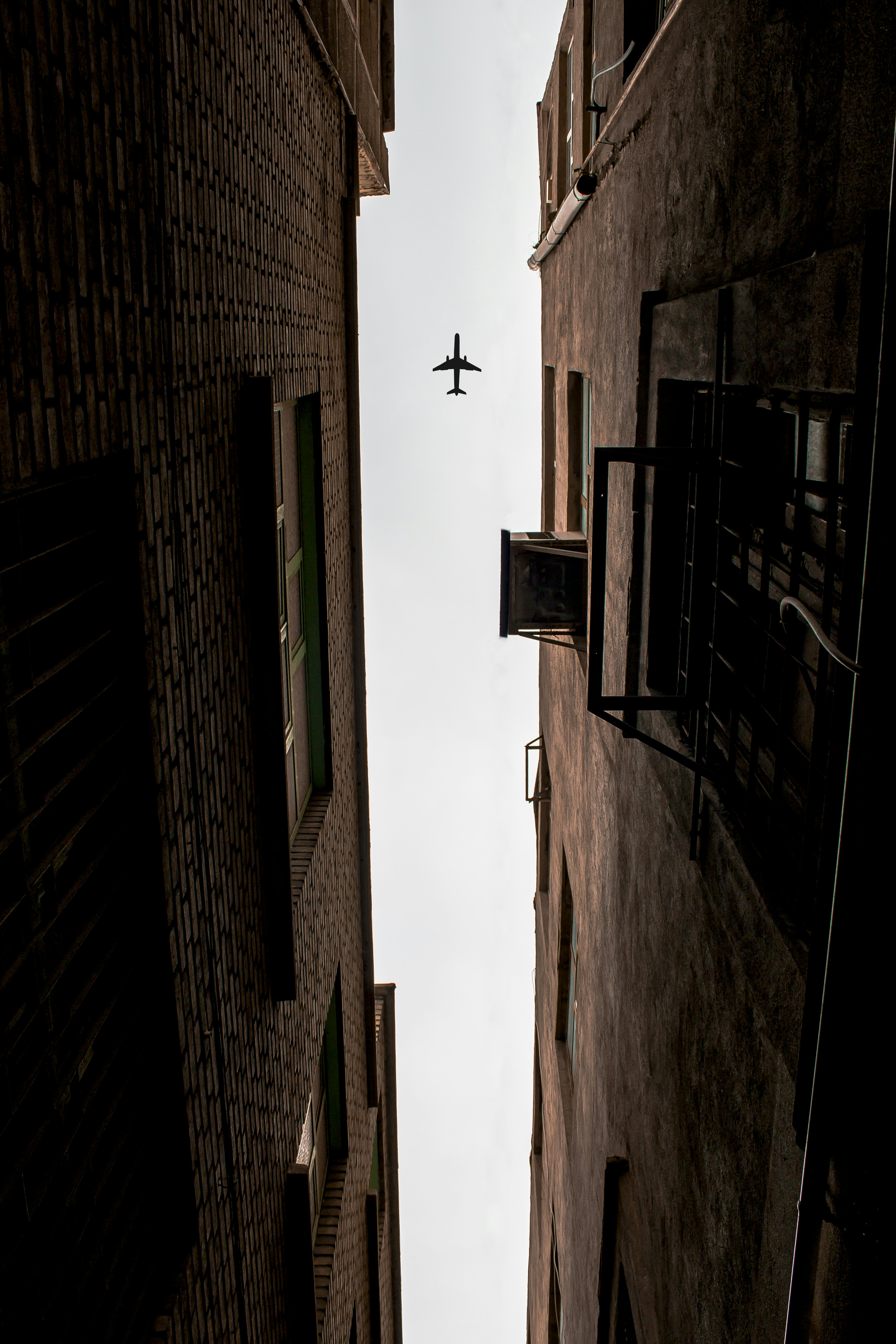 Photograph of a jet silhouetted in a narrow alley framed by tall brick buildings. The pale sky opens above.