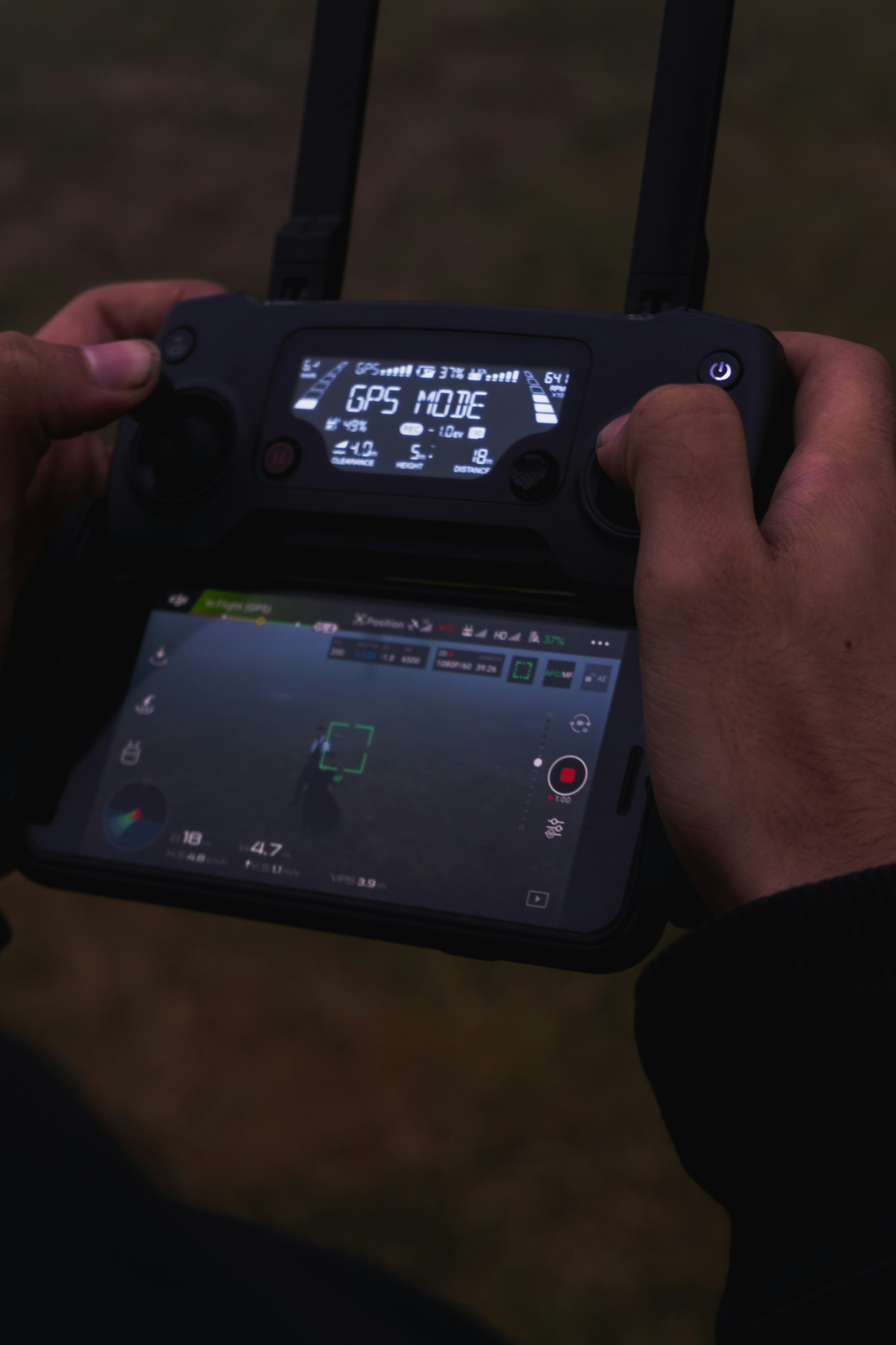 Hands gripping a drone controller displaying GPS mode and flight data on the screen, set against a dimly lit background.