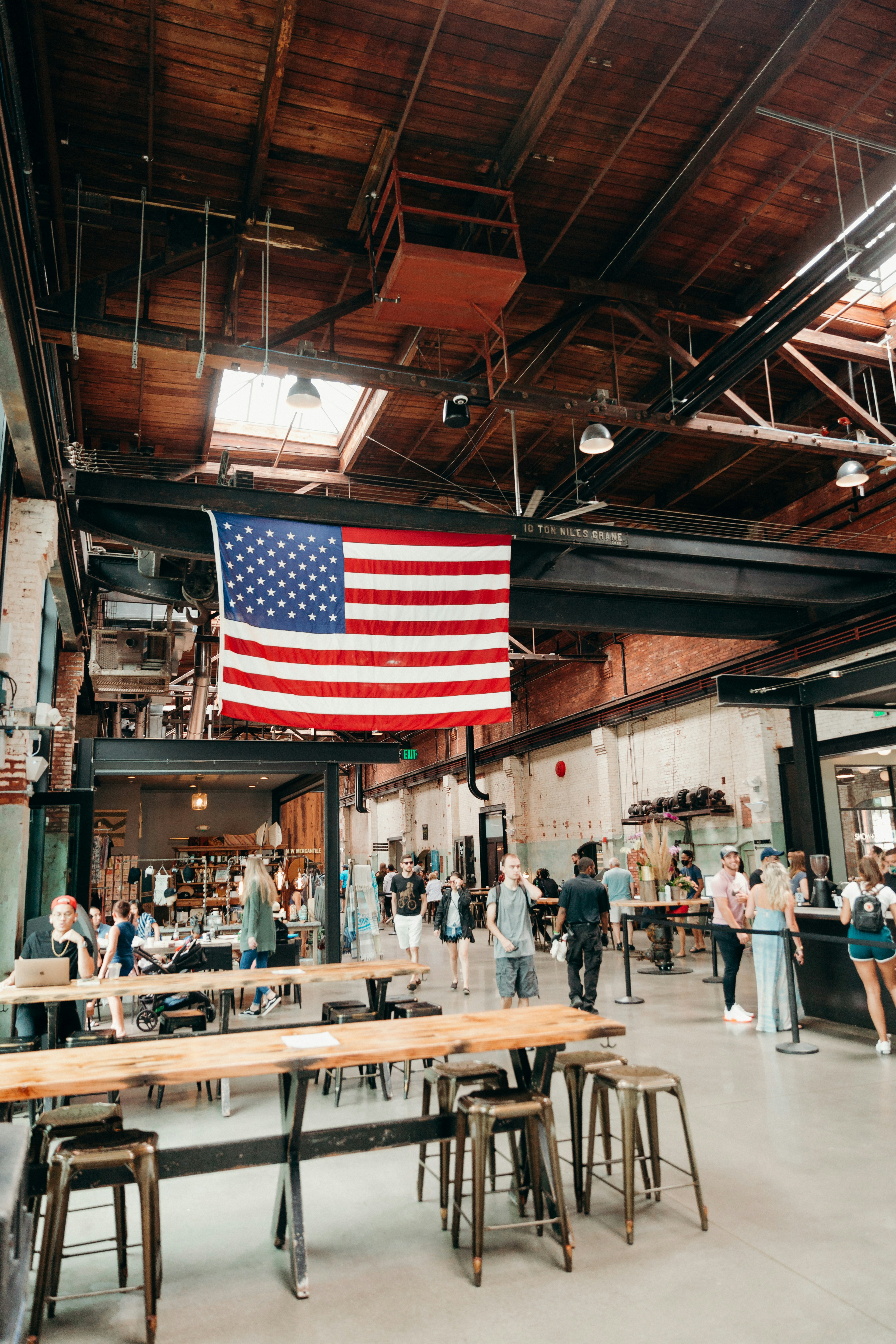 A vibrant indoor space featuring a large American flag, with people enjoying the atmosphere and engaging in conversation. The rustic decor complements the lively setting.