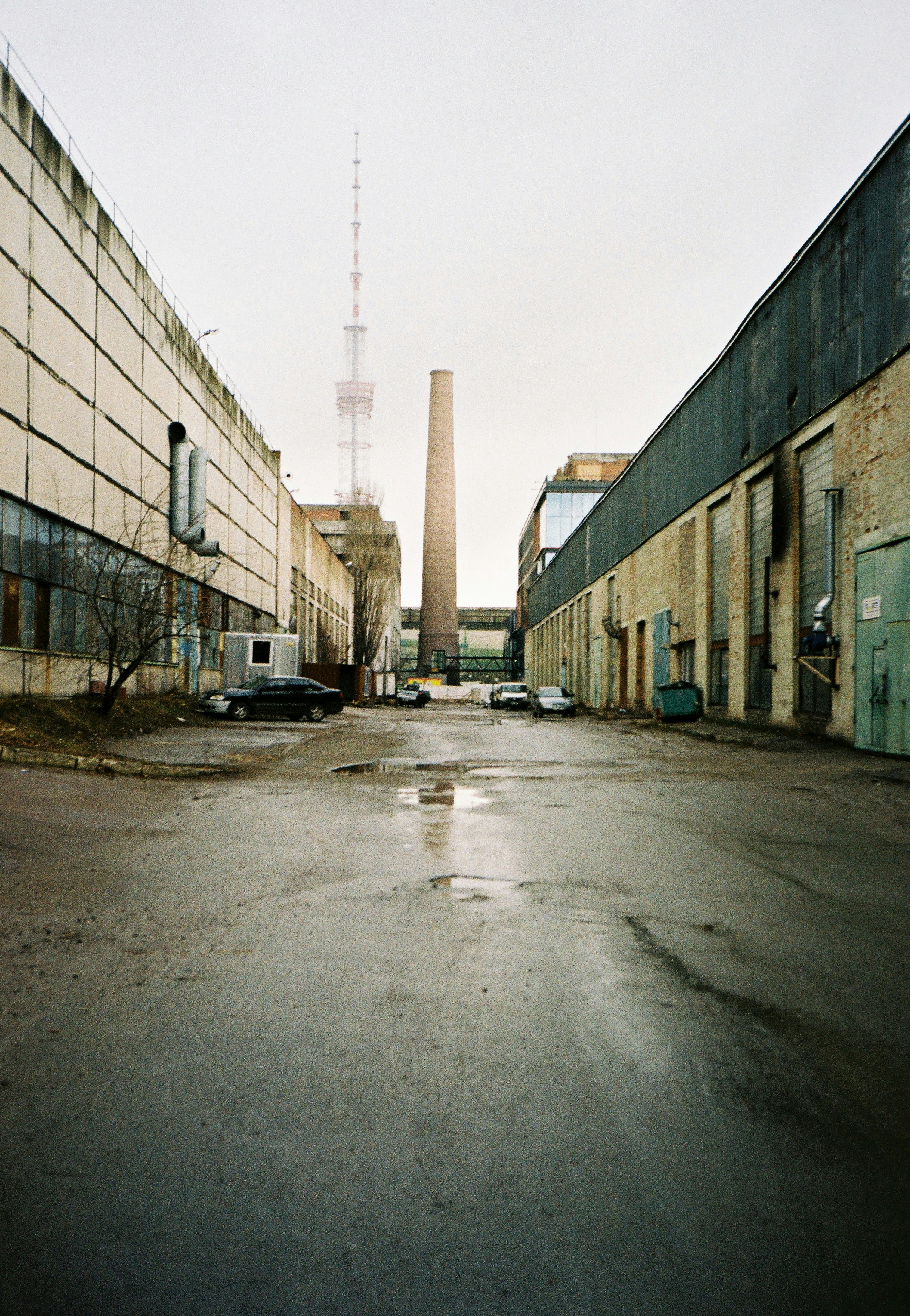 Abandoned industrial alleyway with a towering smokestack and a distant communication tower, reflecting a bygone era of manufacturing.