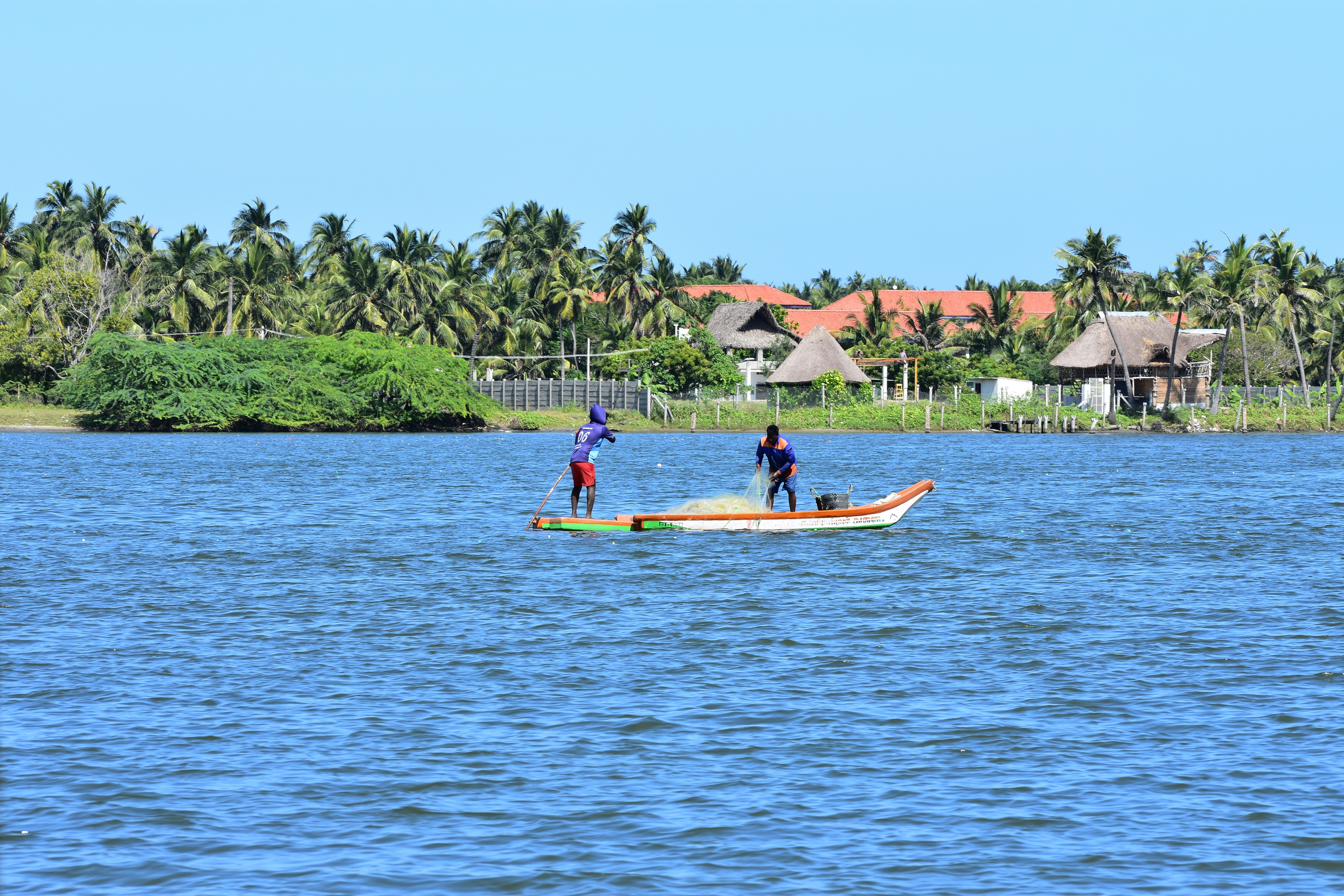 Two fishermen navigate a small boat on calm waters with a backdrop of coconut trees and coastal houses.