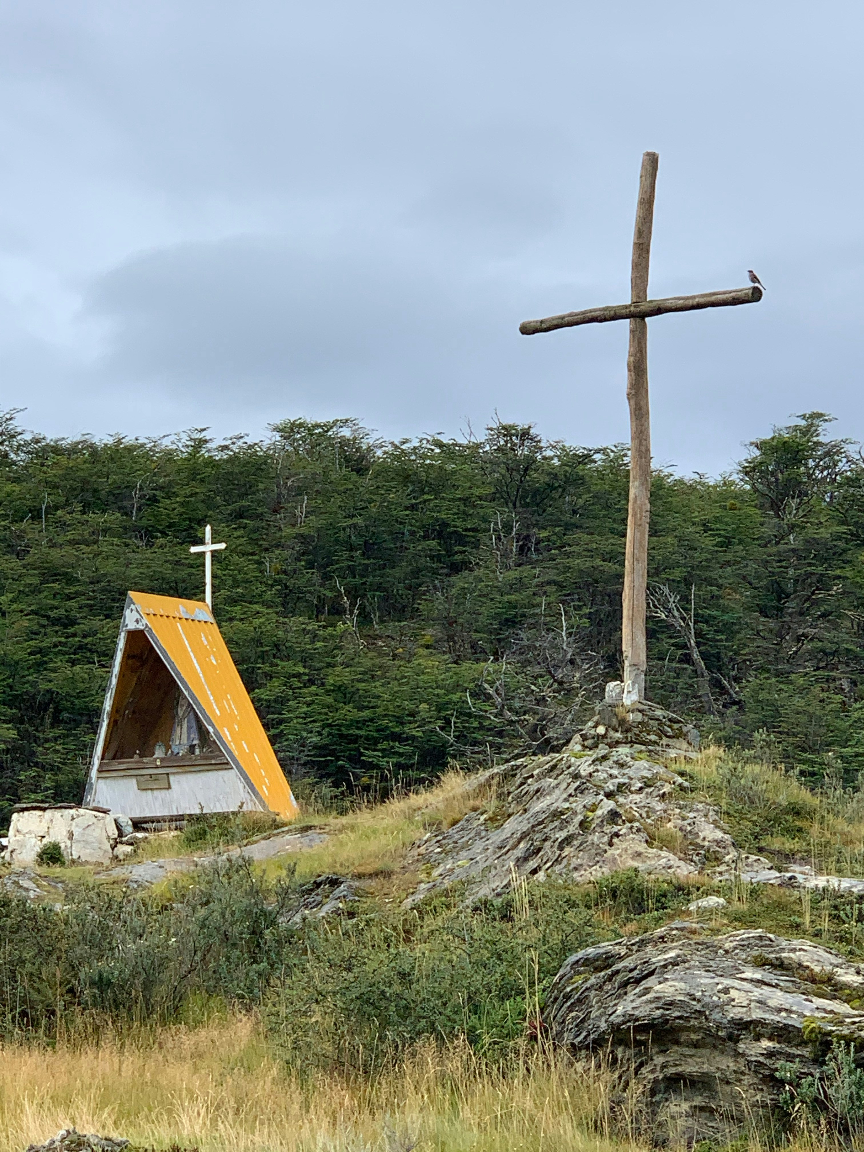 A triangular shelter with a vibrant orange roof stands beside a tall wooden cross, surrounded by lush greenery and rocky terrain.