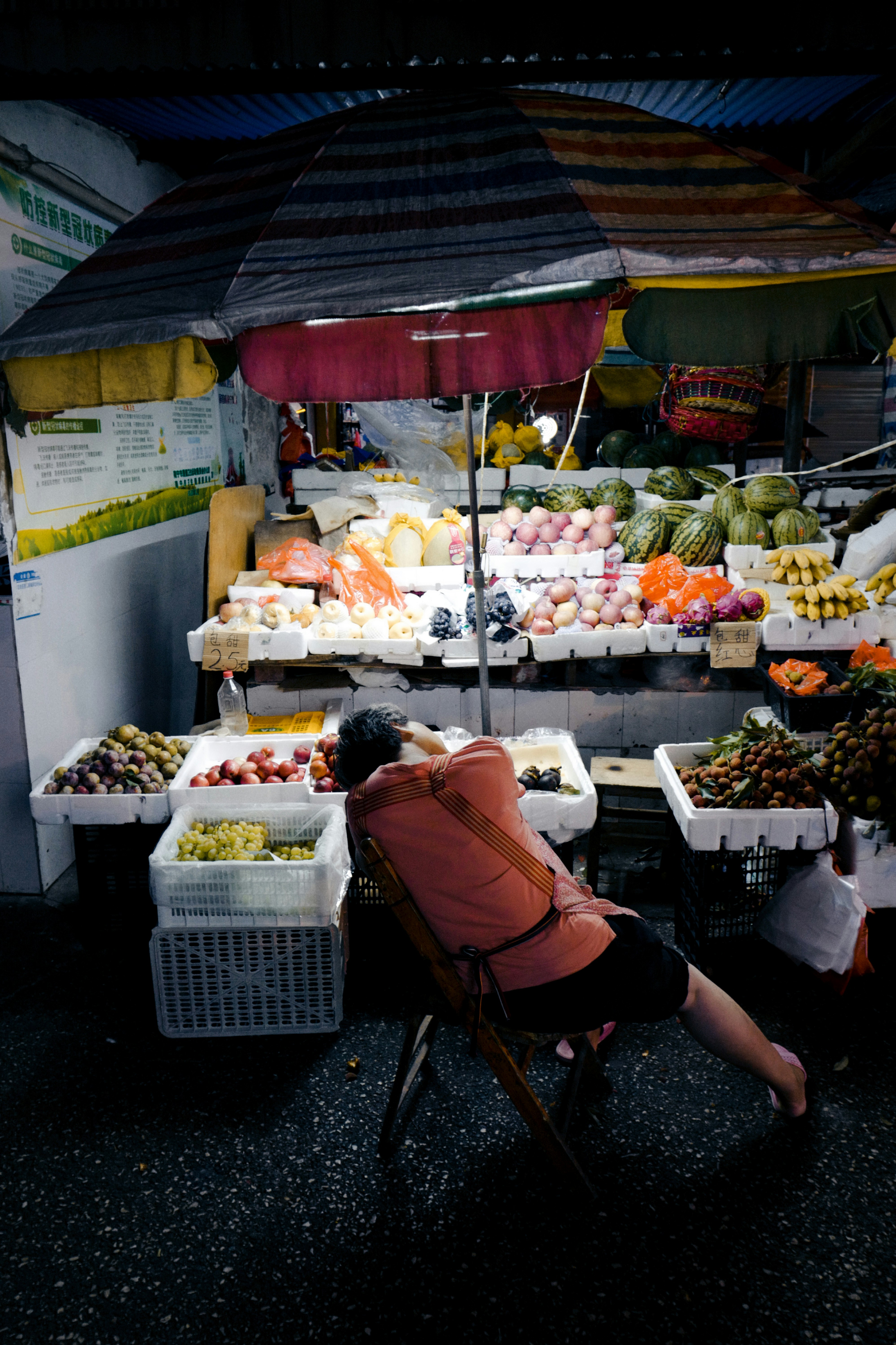 A vendor rests beneath a striped umbrella at a bustling fruit market, surrounded by vibrant displays of fresh produce. The scene captures the contrast between activity and tranquility.