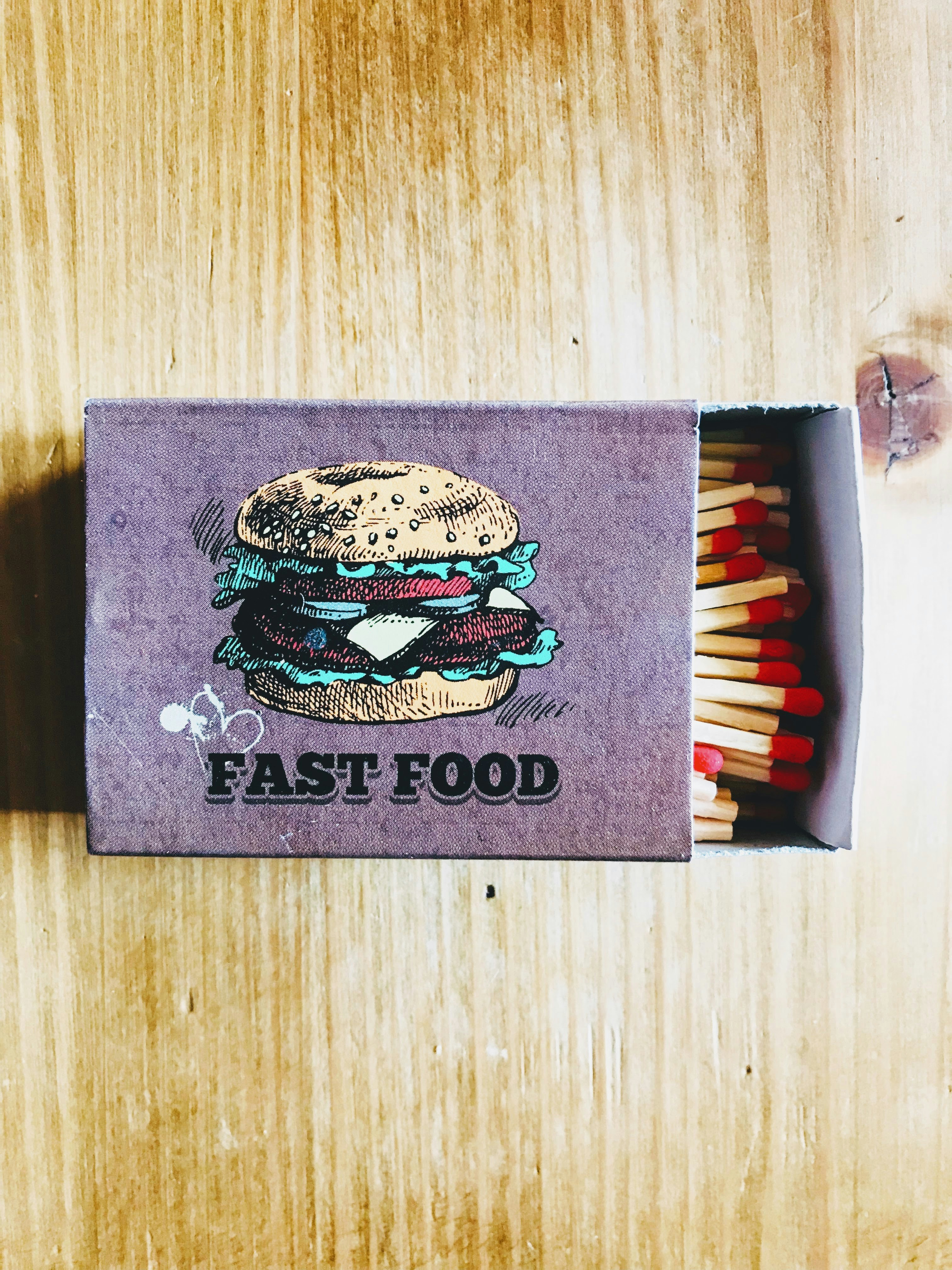 Illustrated matchbox featuring a colorful hamburger design, labeled 'FAST FOOD,' with matches peeking out from one side.