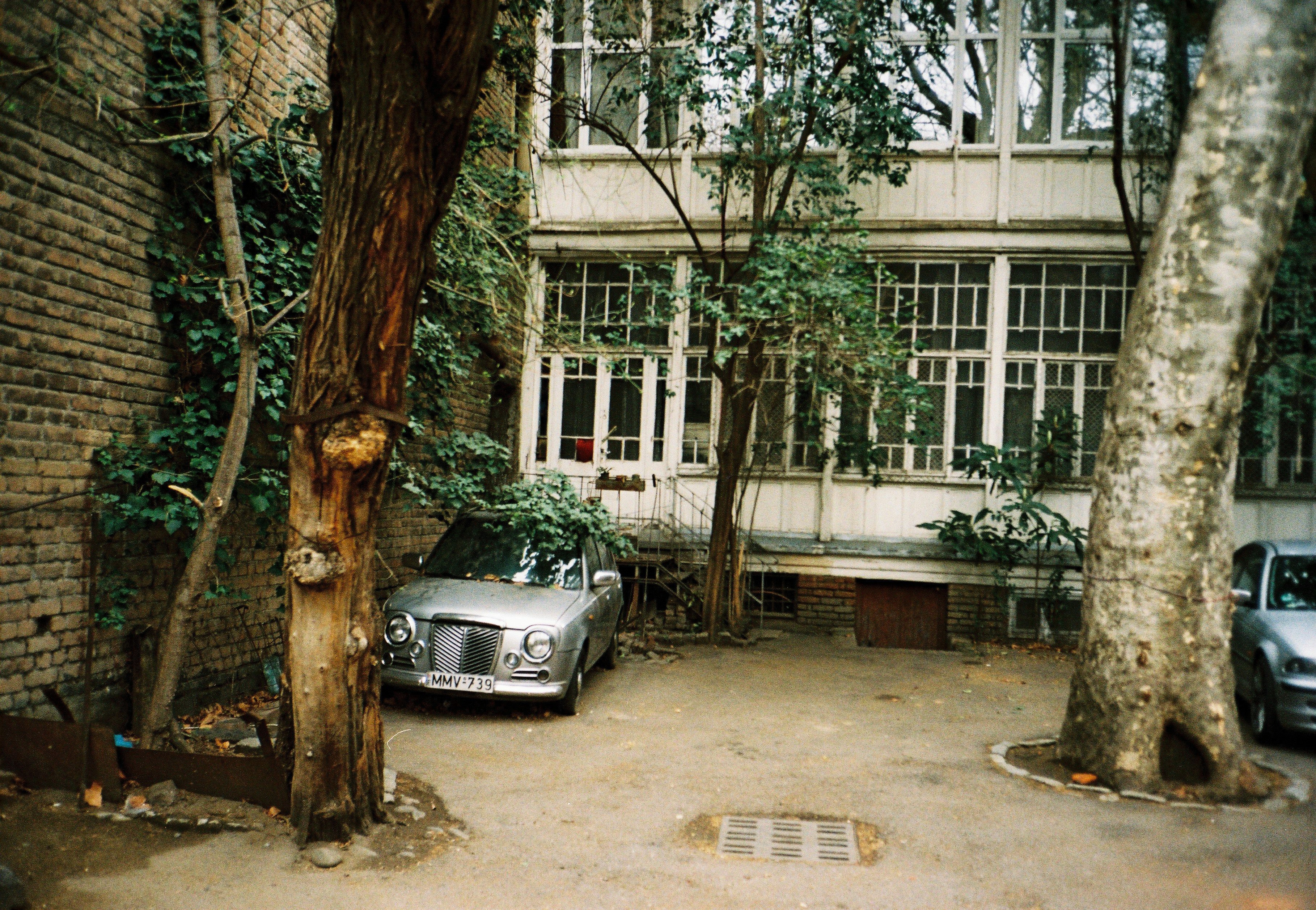 Classic silver car nestled among trees in a quiet courtyard, framed by a rustic brick wall and large windows. 