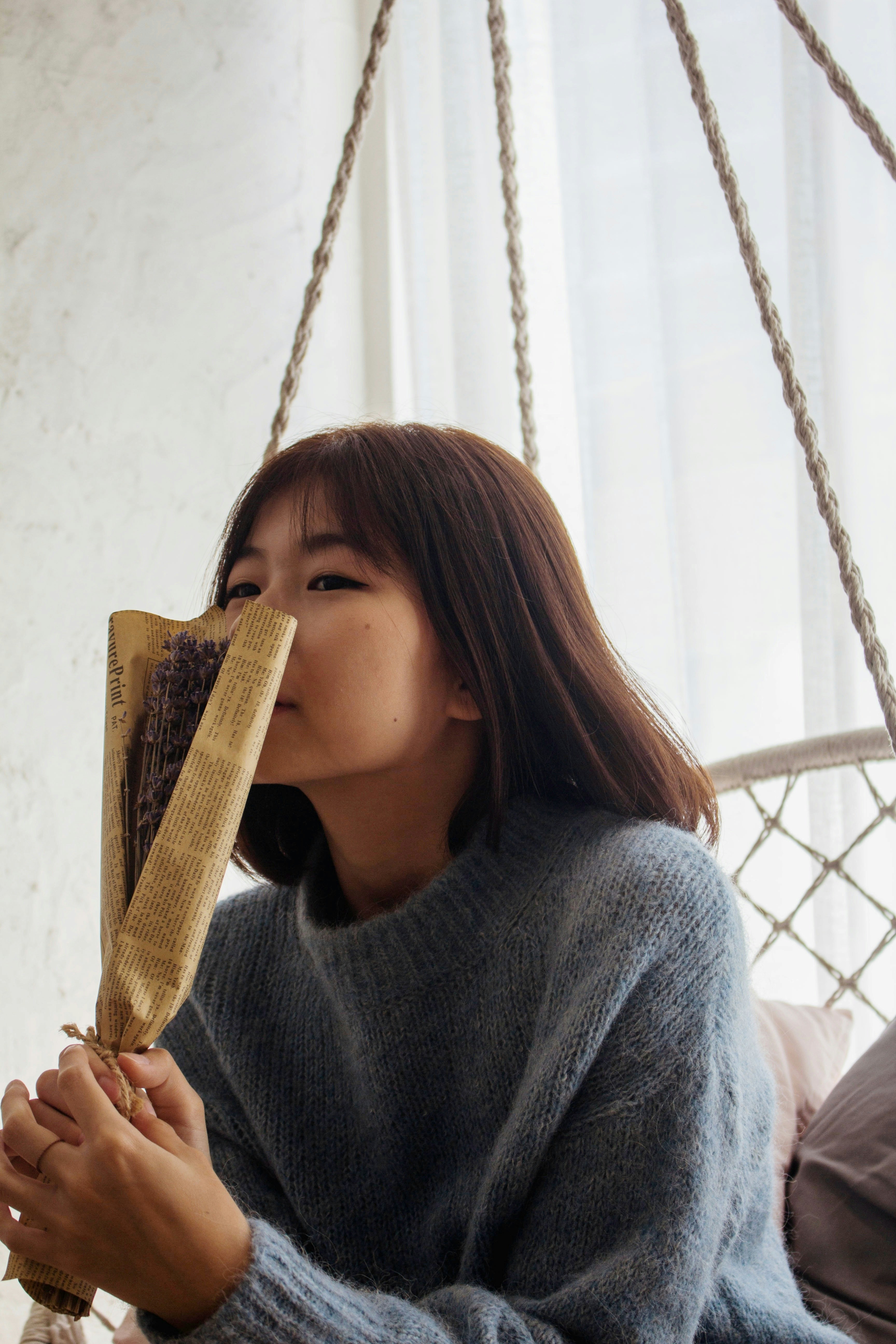 A young woman gently holds a bouquet of lavender, her expression reflecting tranquility. Soft natural light filters through the window, enhancing the cozy atmosphere.
