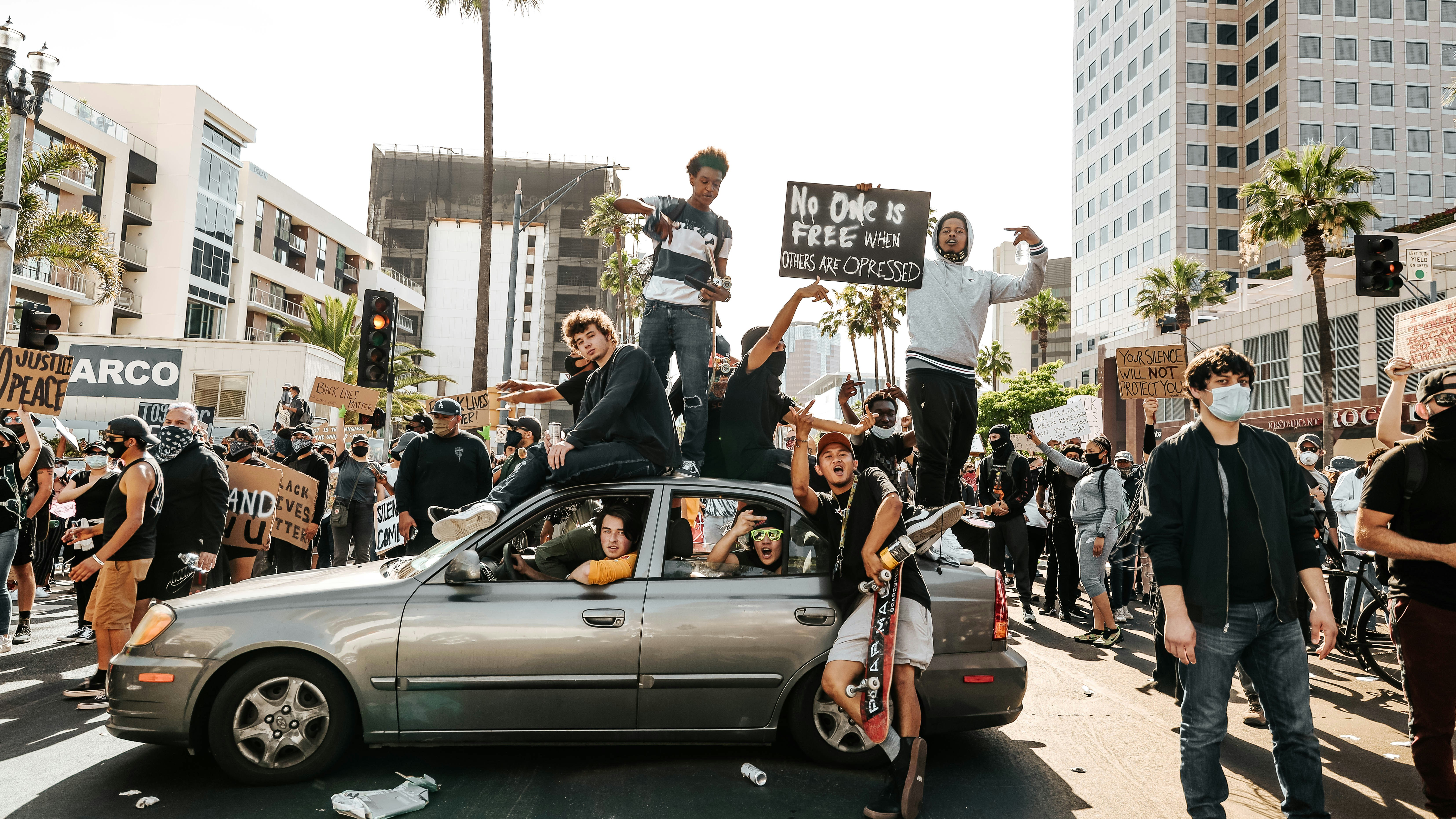 Group gathered around a car holding signs during a daytime protest in a city street.