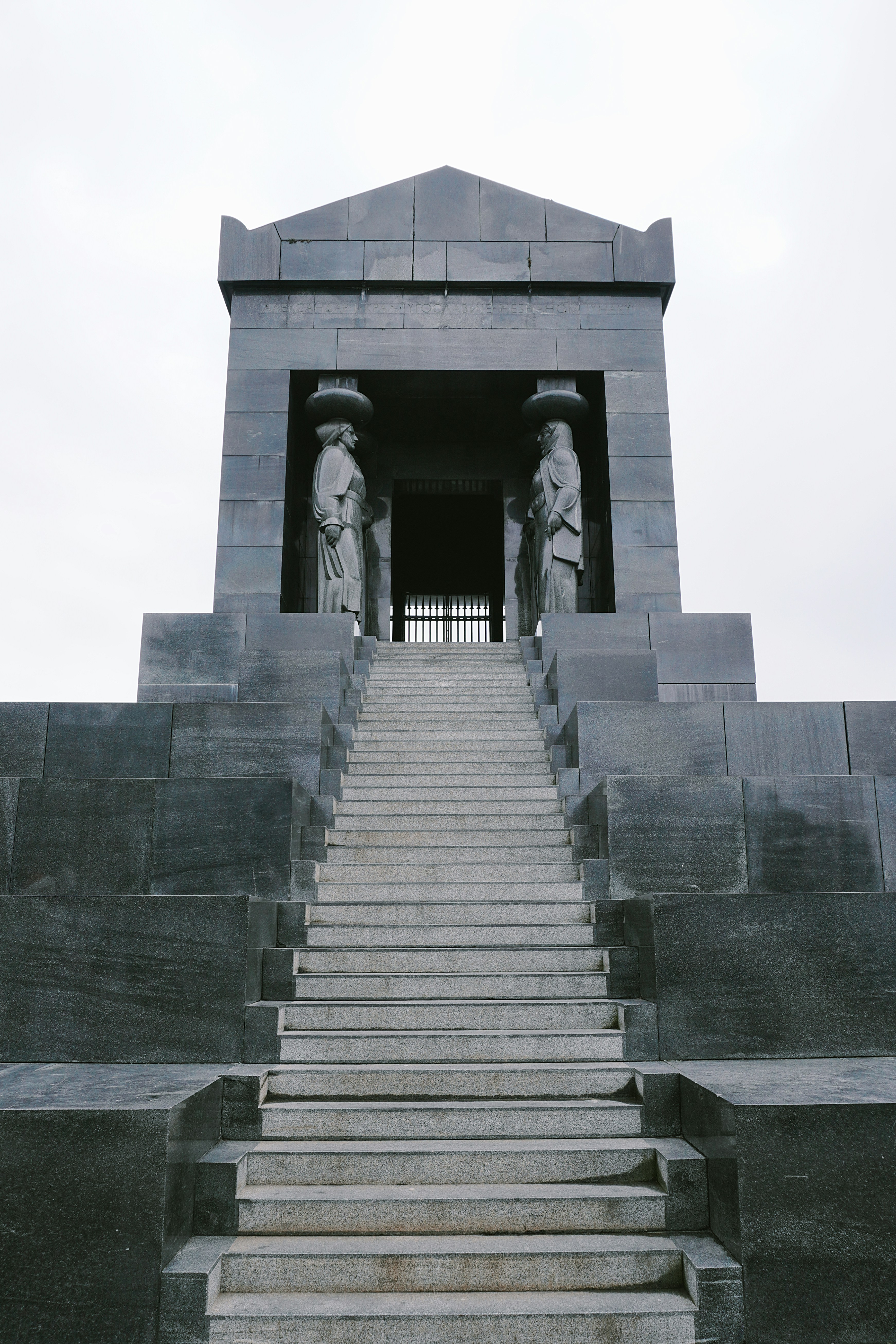 Imposing monument with two statues flanking a grand staircase leading to a temple structure, set against a cloudy sky.
