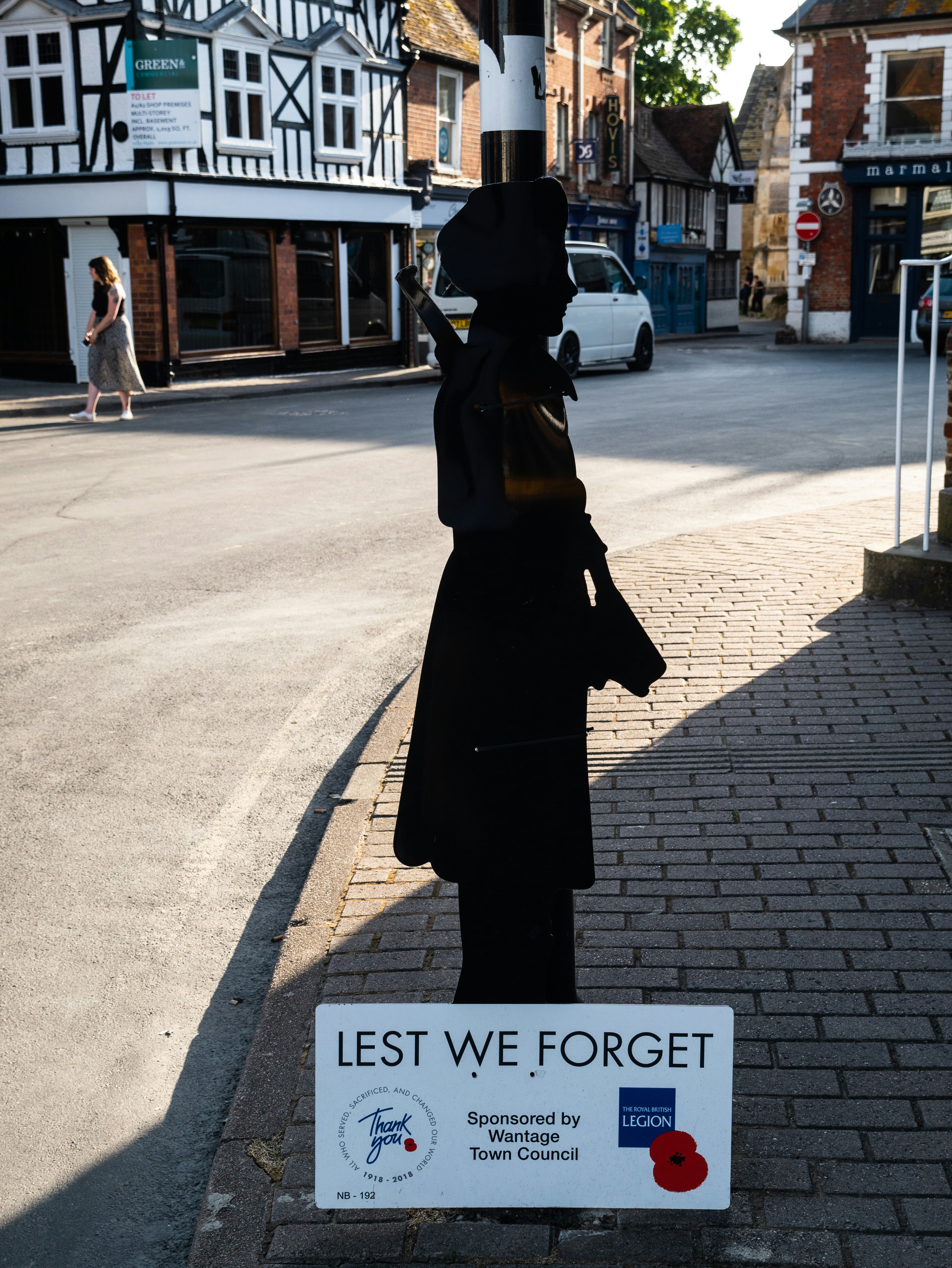 Silhouette of a soldier standing beside a memorial sign reading 'LEST WE FORGET' in a quaint town square.