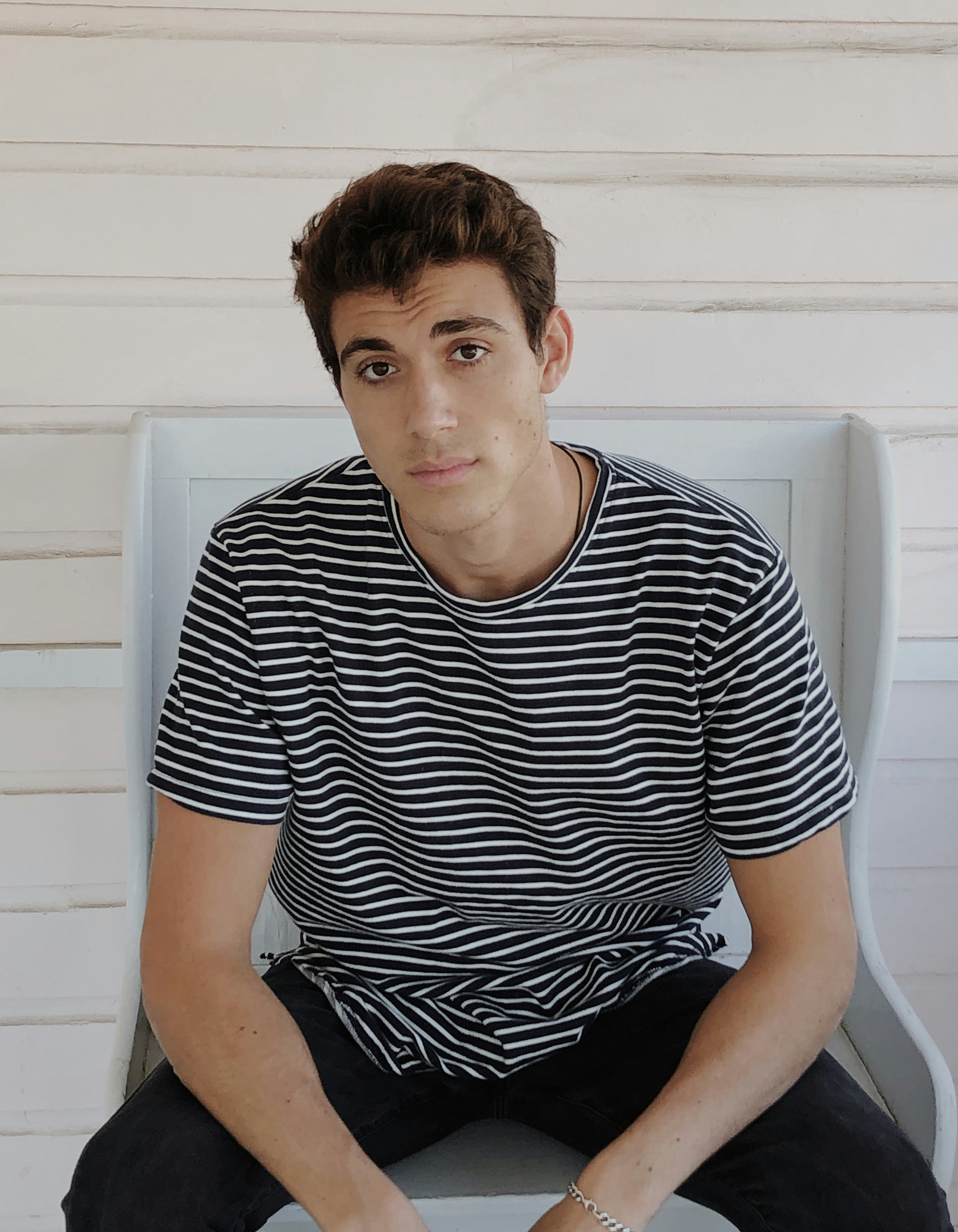 Young man seated on a chair, wearing a striped shirt, gazing thoughtfully at the camera against a light wooden backdrop.