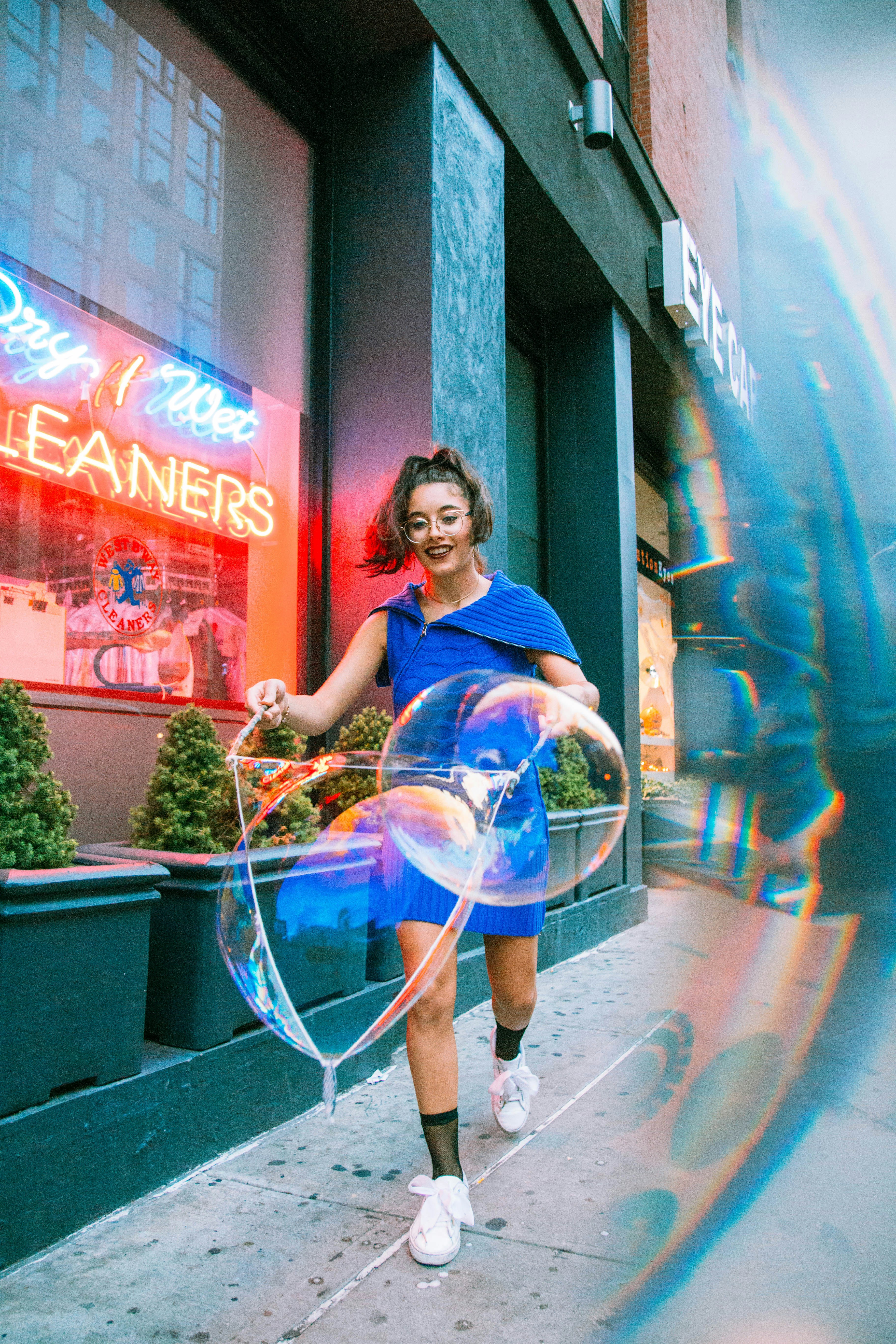 Bubble Waltz Under Neon: Blue Dress and Iridescent Orbs on a City Walk