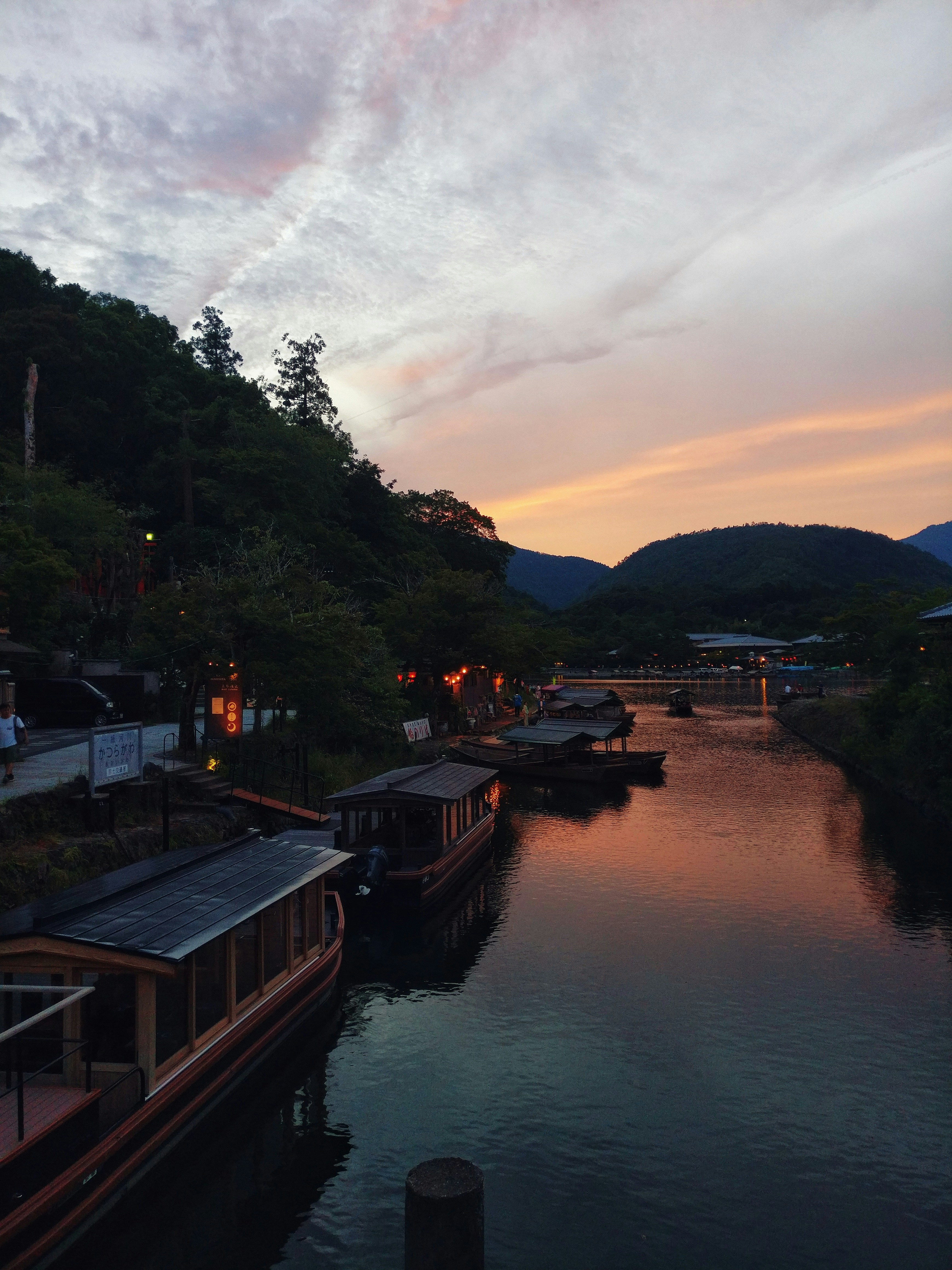 Twilight over a tranquil river village with boats docked along the bank. Lanterns glow along the shore as the peach-pink sky fades.