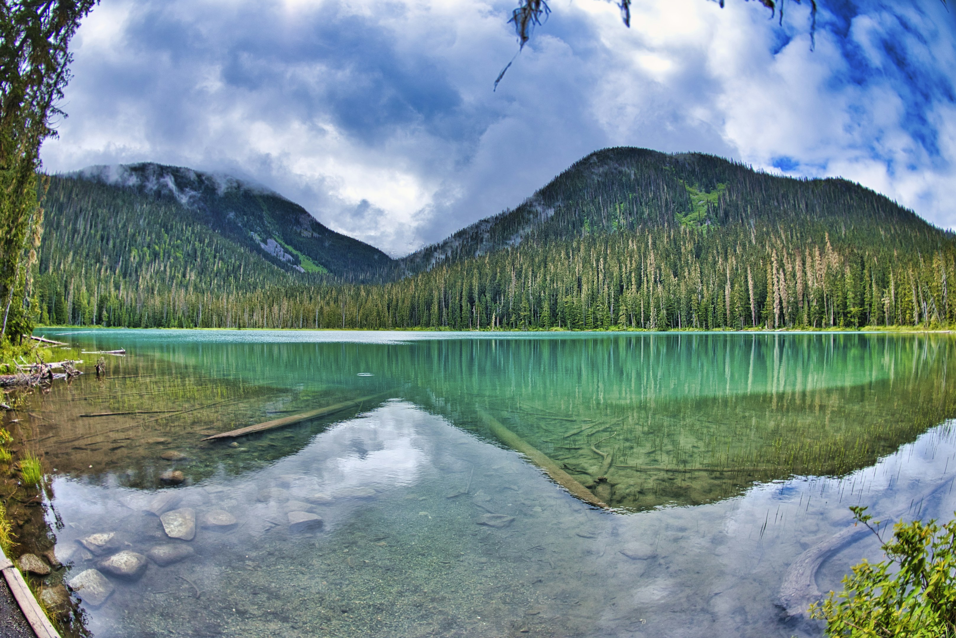 Vibrant turquoise lake reflecting dense evergreen forests and looming mountains under a dramatic sky.