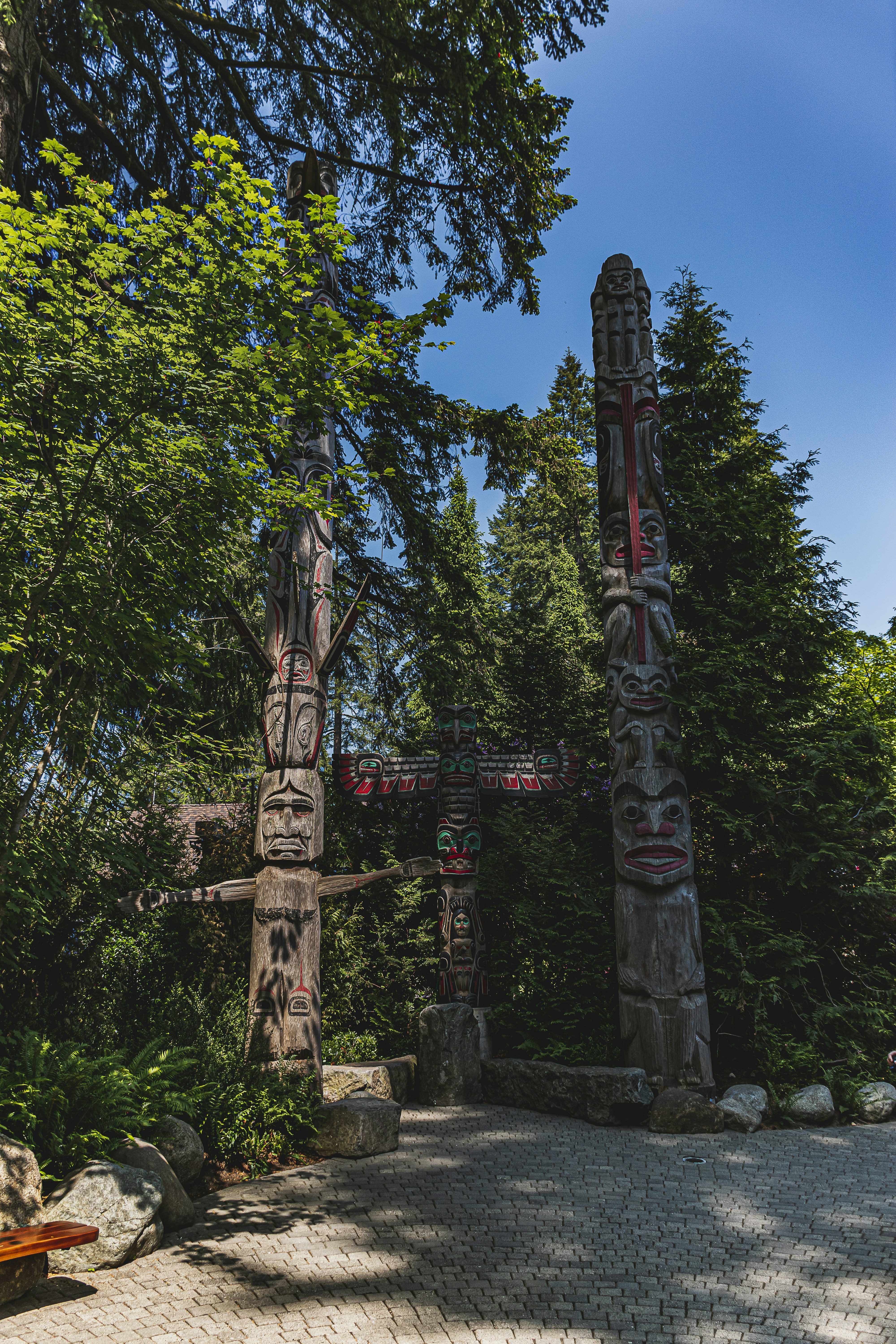 Three intricately carved totem poles rise amidst lush greenery, showcasing vibrant colors and cultural significance. Sunlight filters through the trees, highlighting the artistry.