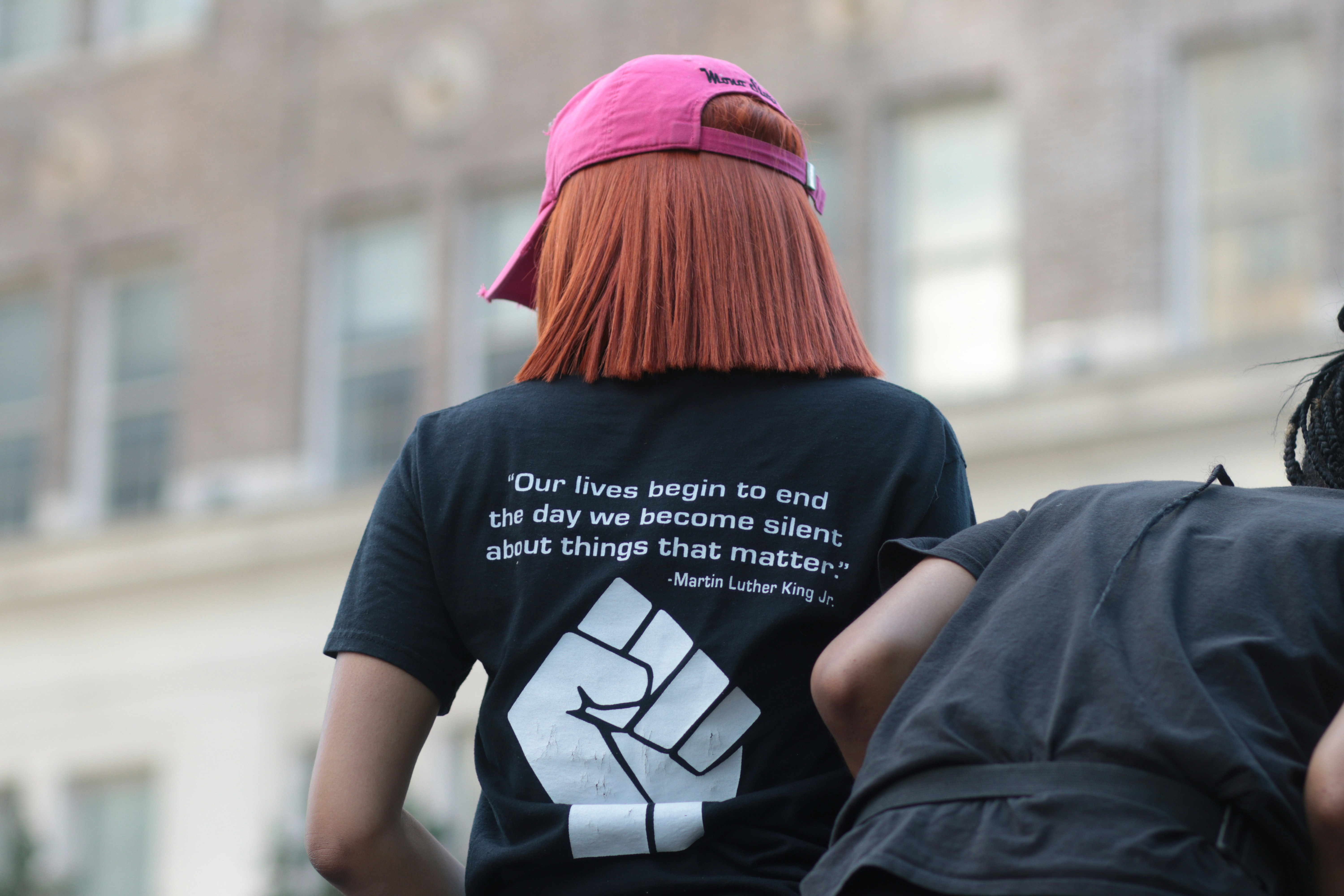 Person in a red cap and black shirt with a raised fist symbol stands against an urban backdrop.