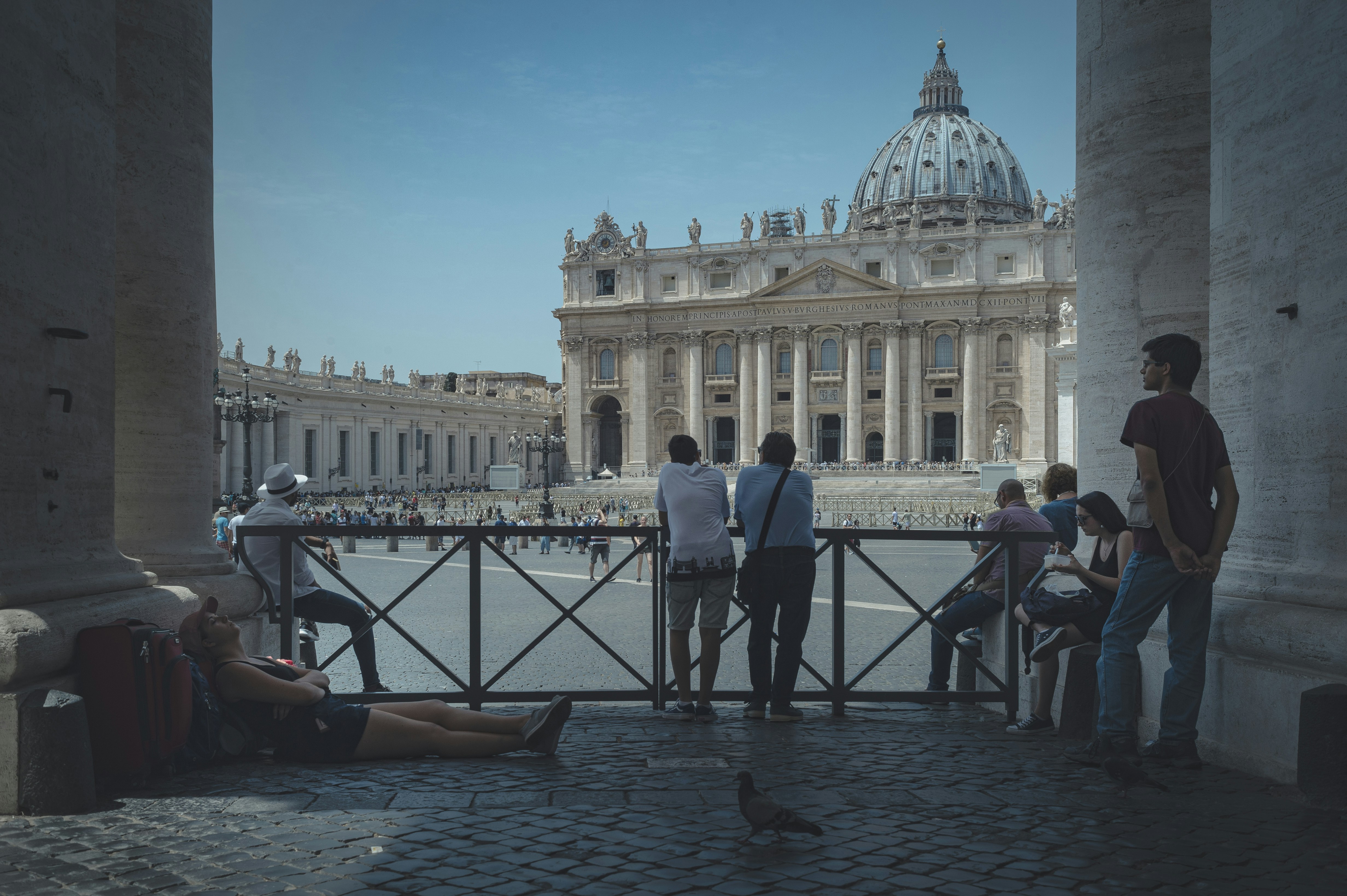Visitors gather under an archway with St. Peter's Basilica in the background on a sunny day.