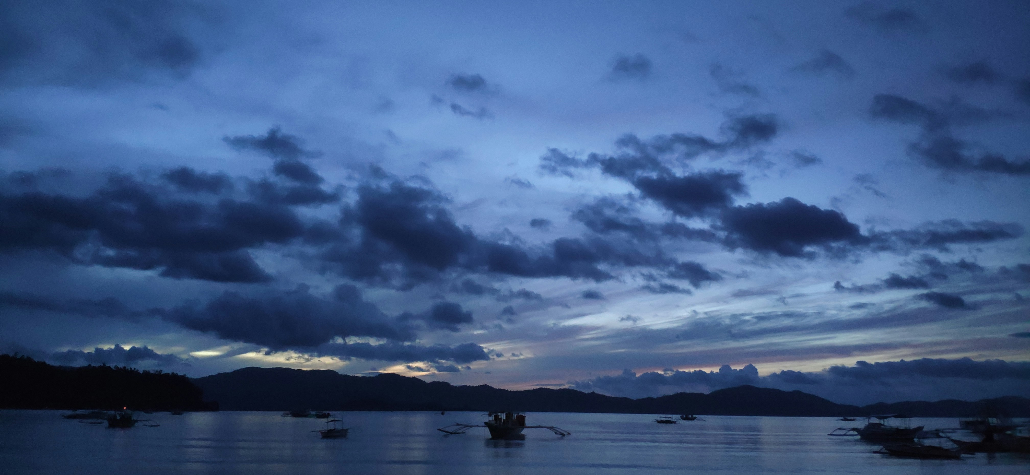 Cloud-dappled sky at dusk over a tranquil sea, silhouetted by distant mountains.