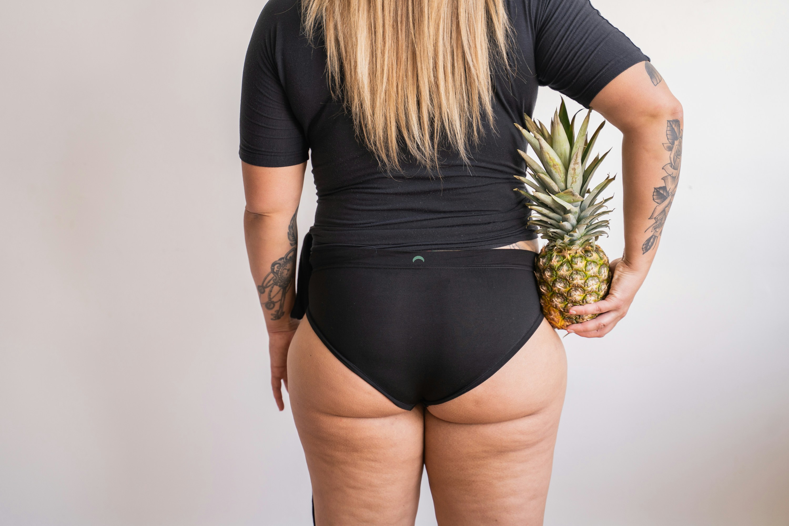 Model showcasing a playful pose with a pineapple tucked behind in a stylish black outfit. The image highlights a unique blend of fashion and humor.