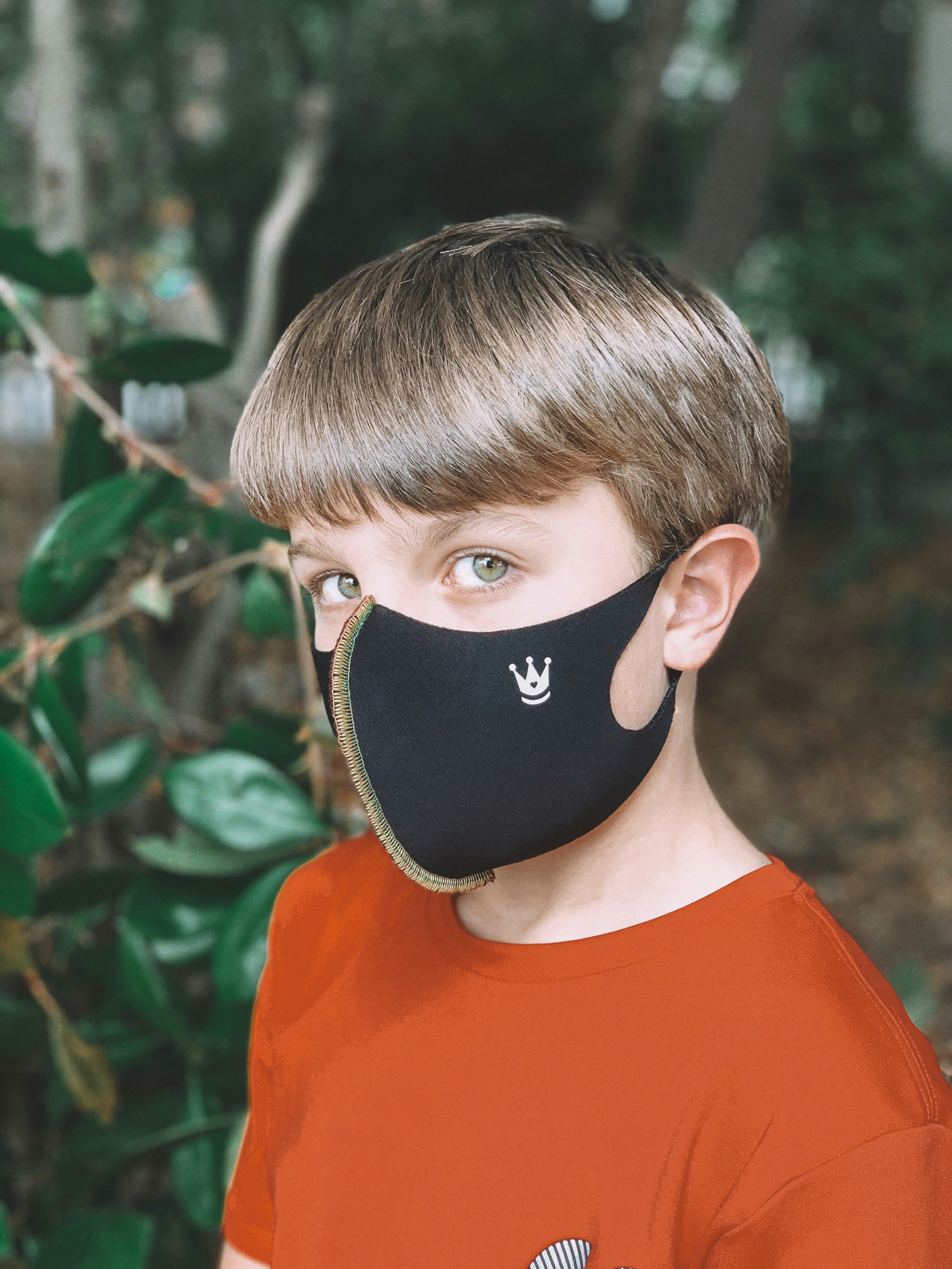 Child wearing a black mask with a crown logo, surrounded by lush greenery. The image captures a moment of youthful resilience amidst changing times.