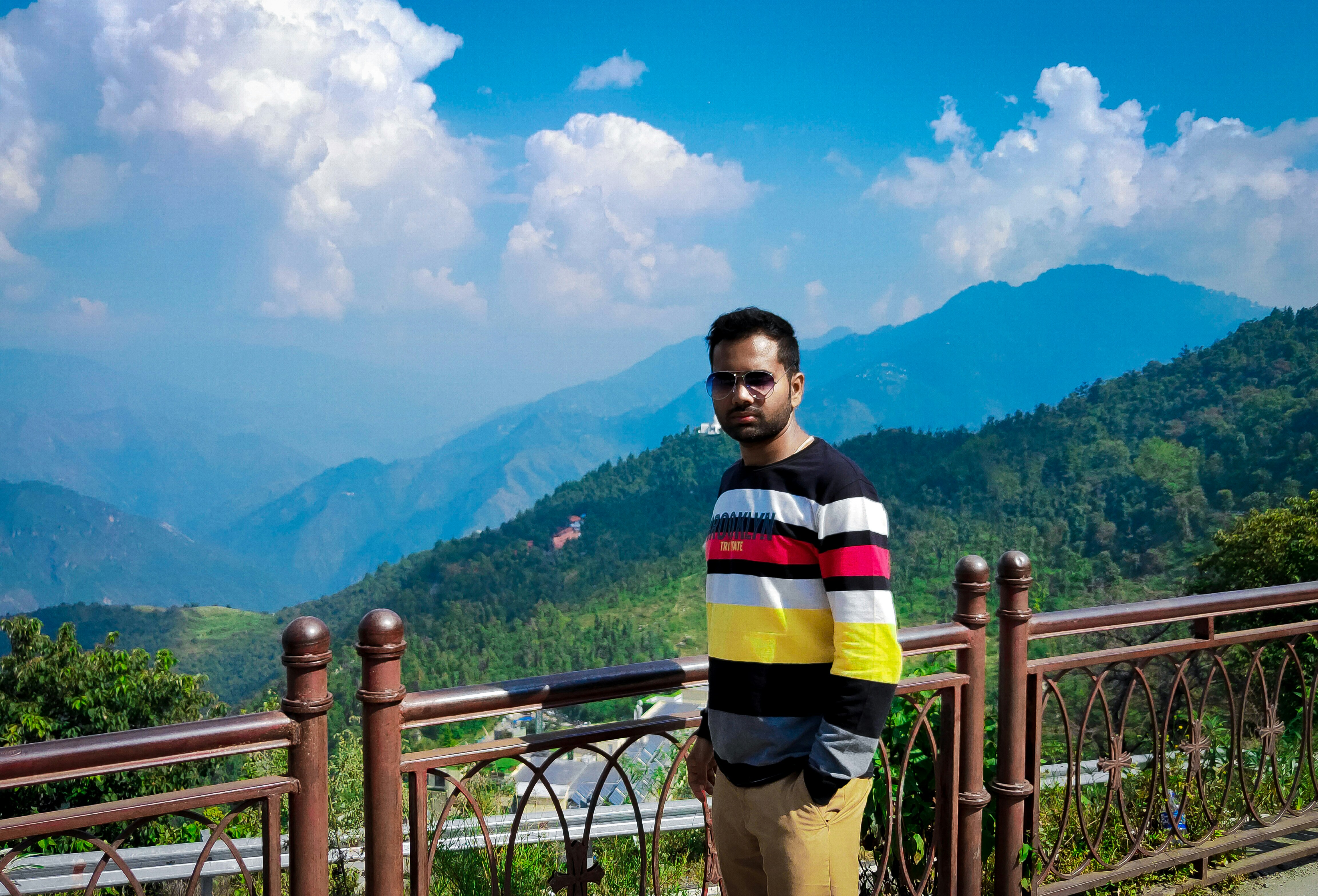 Person standing against a backdrop of rolling mountains and a clear blue sky, with a fence in the foreground. The scene captures a tranquil moment in nature.